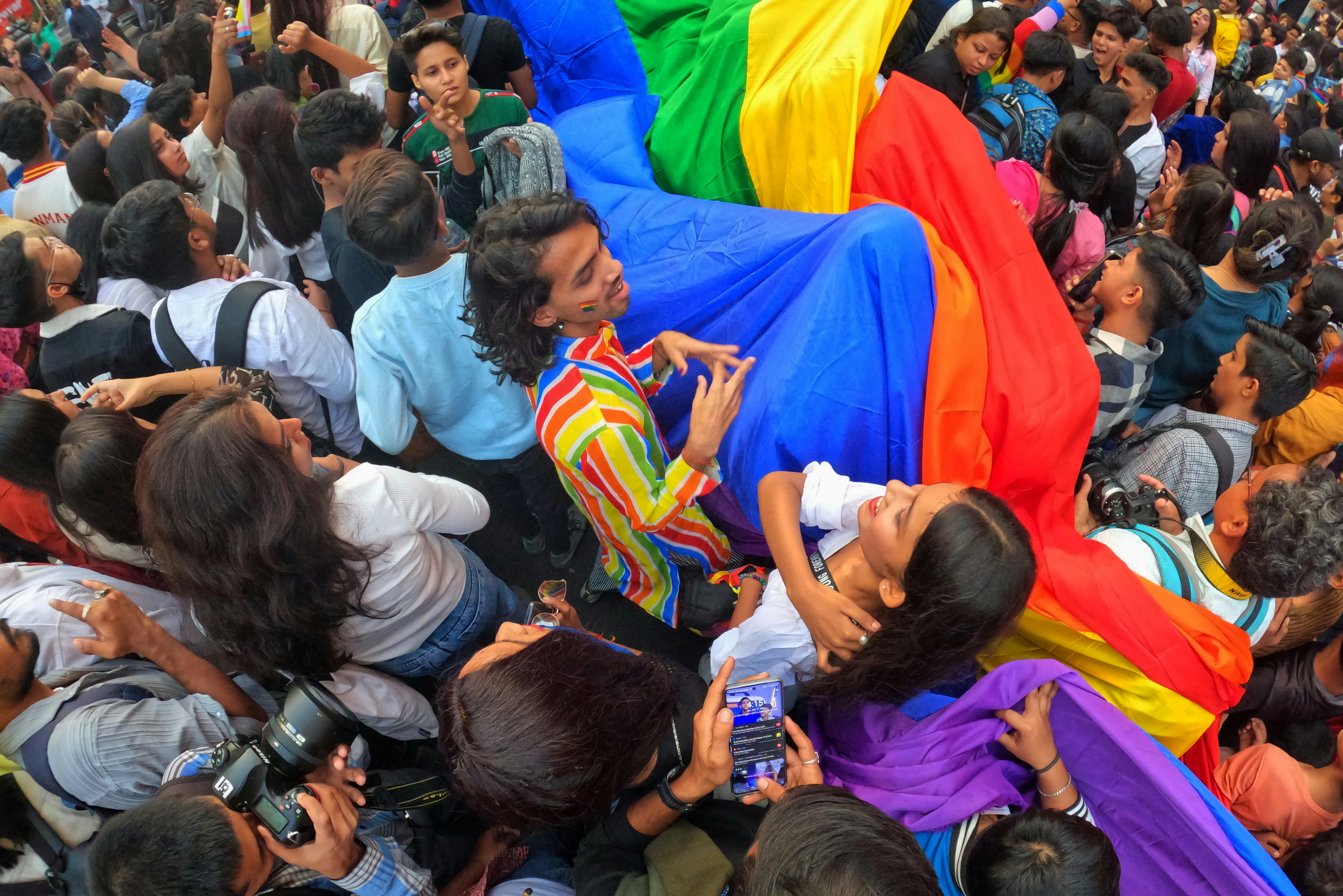Crowd gathered under a large rainbow flag, celebrating diversity with joyful expressions.