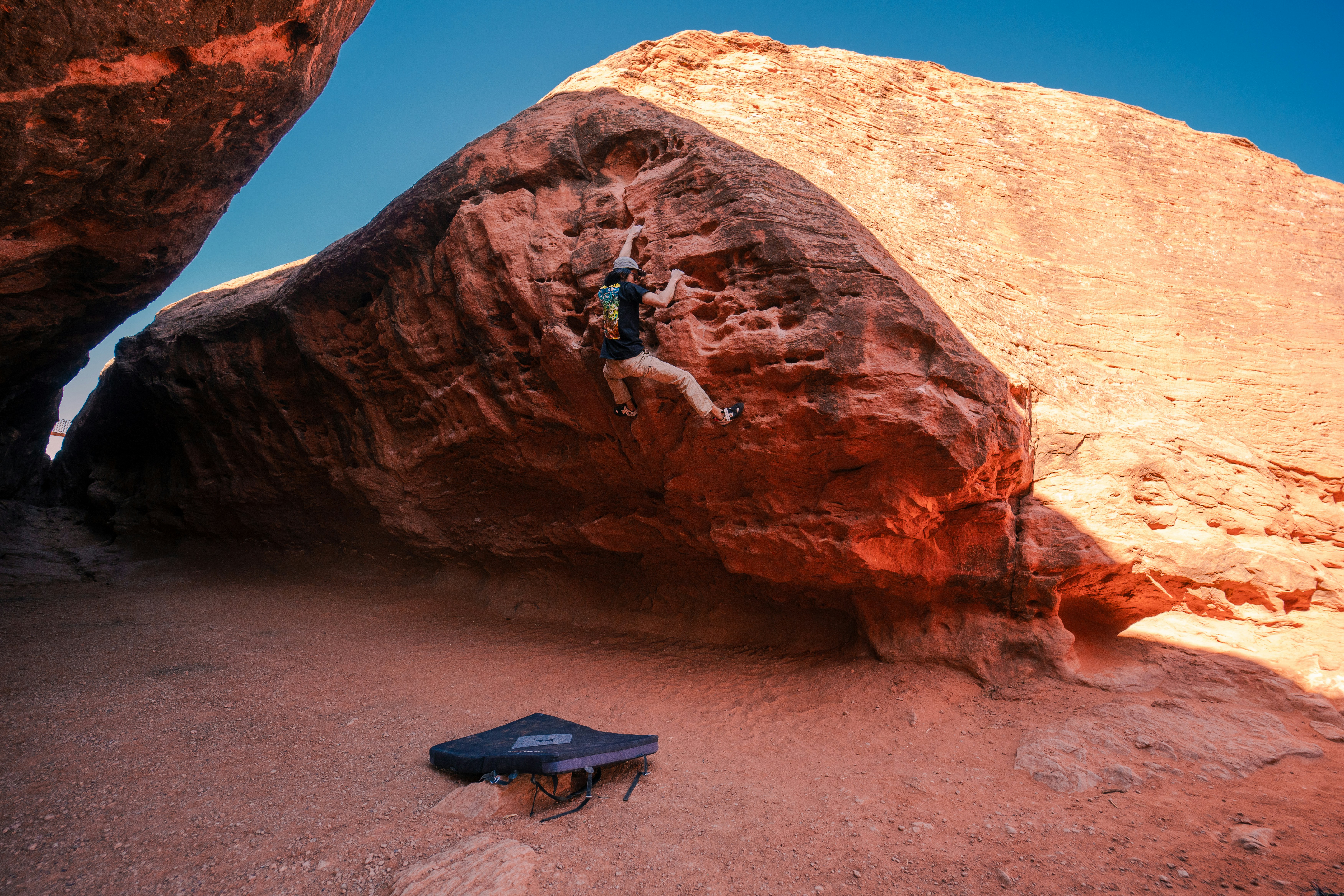 Rock climber scaling a rugged, sunlit sandstone boulder under a clear blue sky.