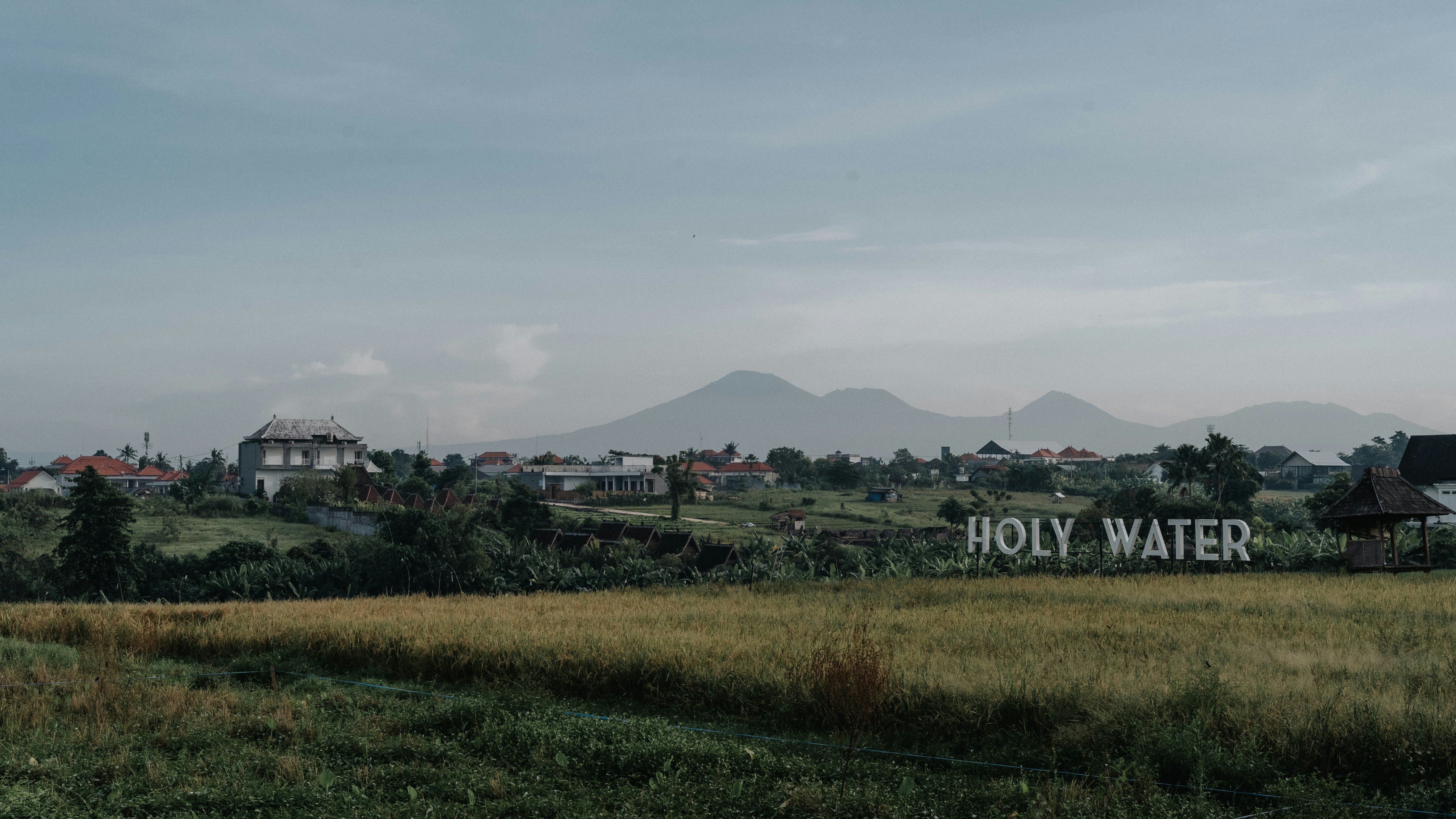 Expansive green fields with 'HOLY WATER' sign, set against distant mountains under a cloudy sky.
