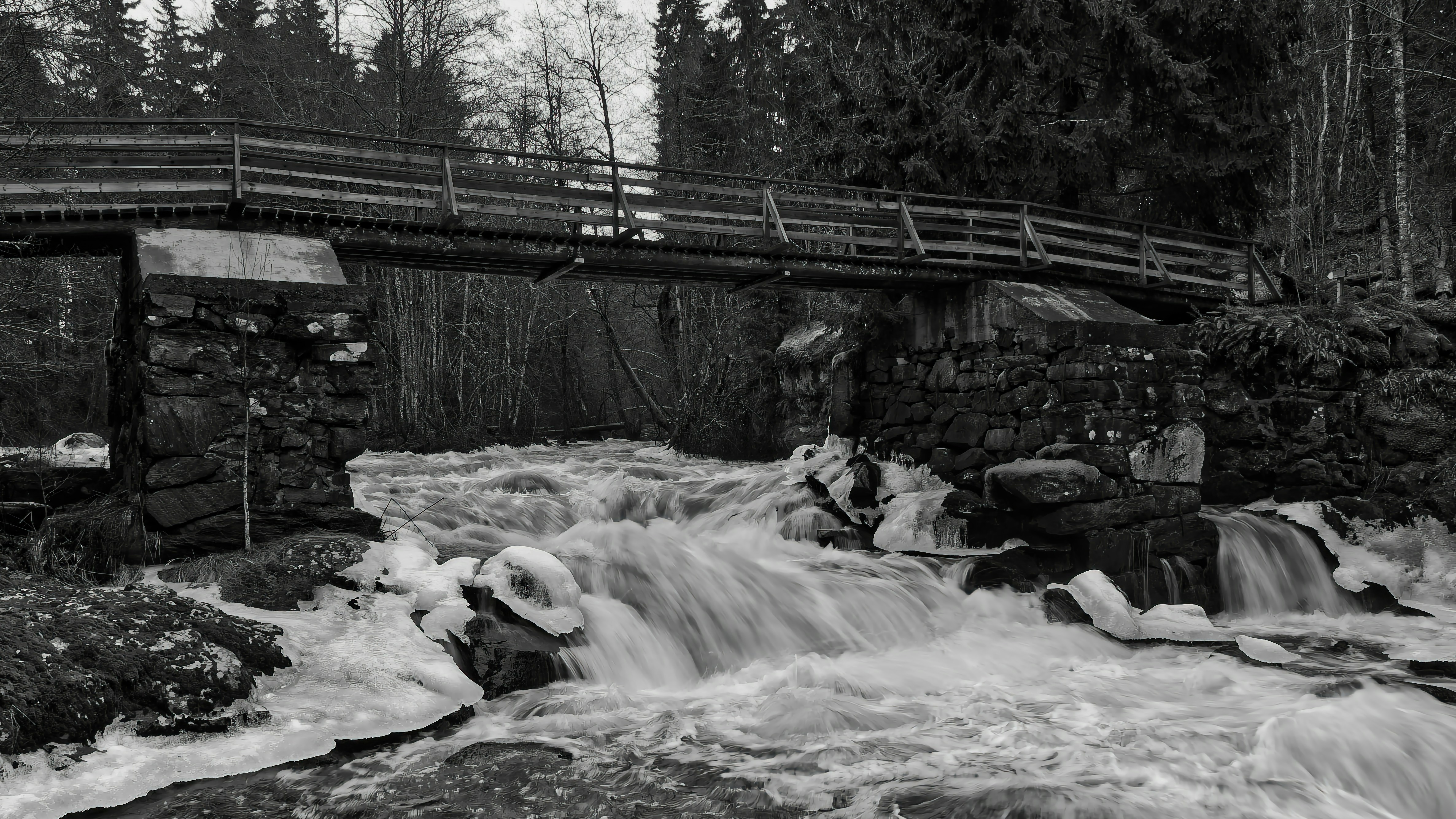 Wooden bridge spanning a frothy river in a forested landscape during winter.