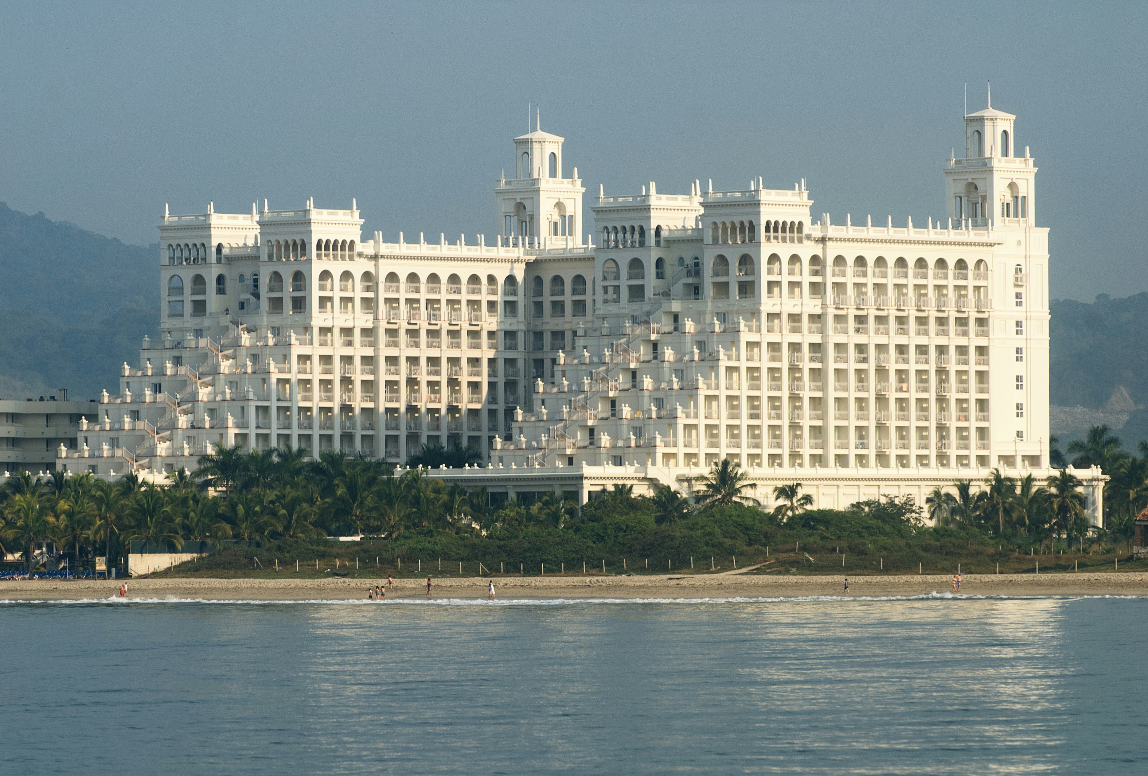 Grand white hotel with tiered balconies set against a tropical beach backdrop.