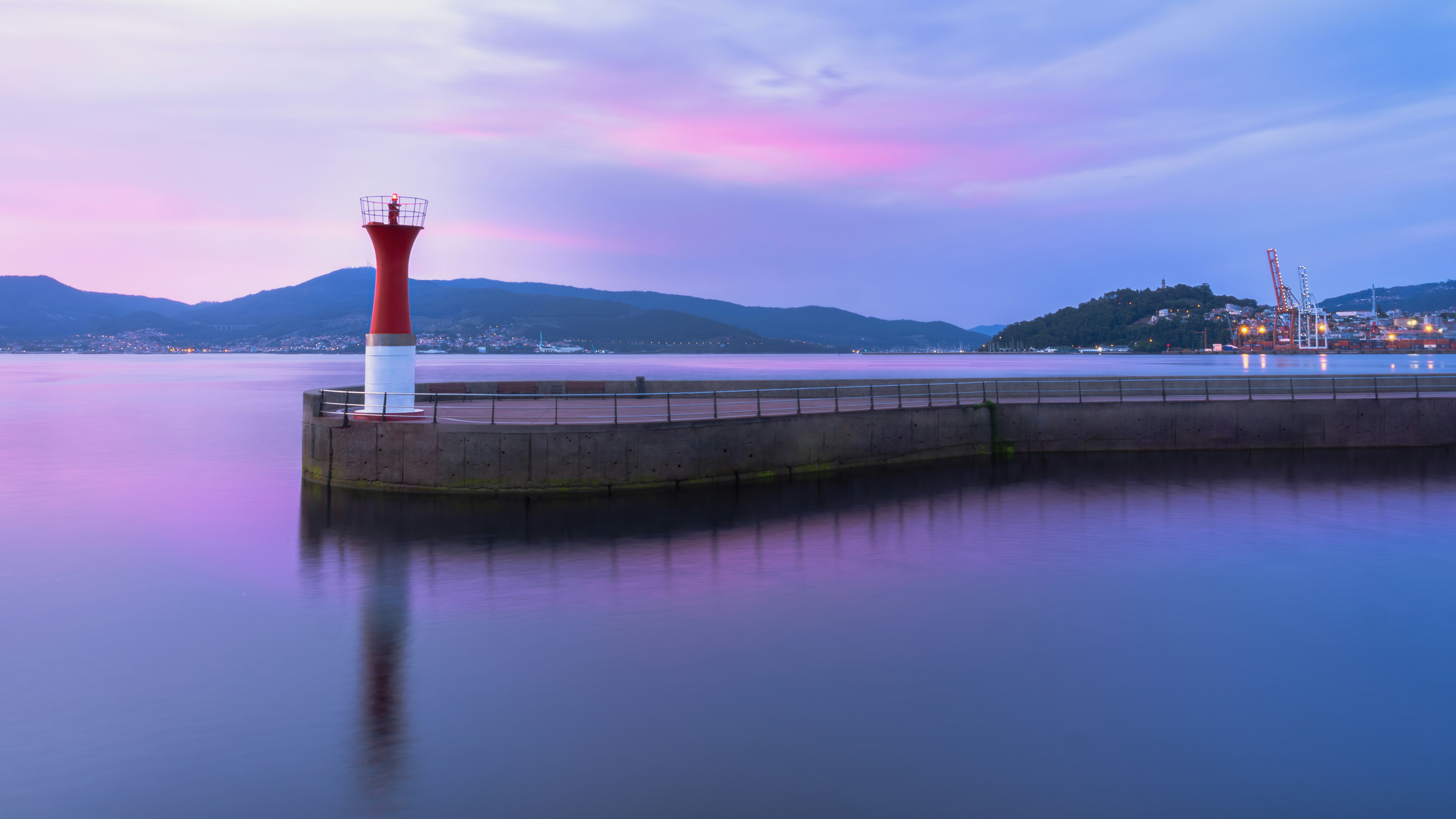 Red and white lighthouse on a calm harbor reflects in tranquil evening waters with a pink and blue sky.