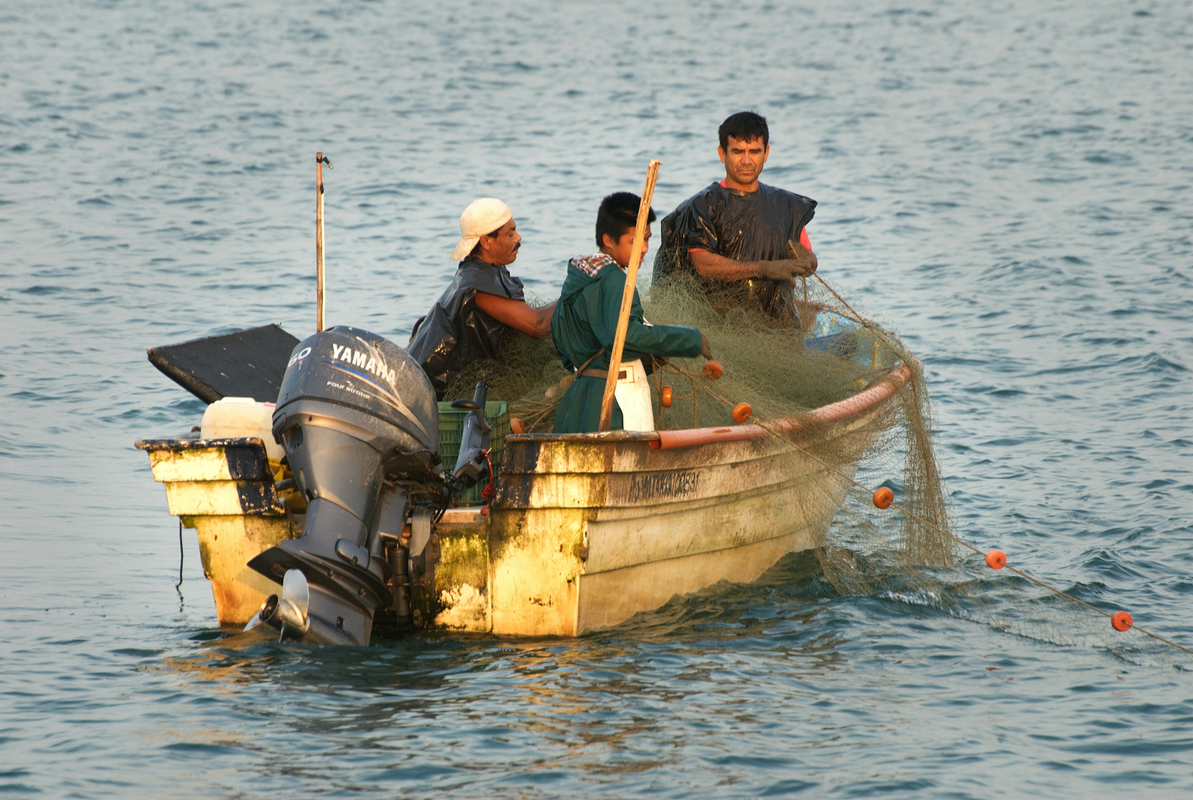 Three fishermen hauling nets on a small motorboat under soft morning light.