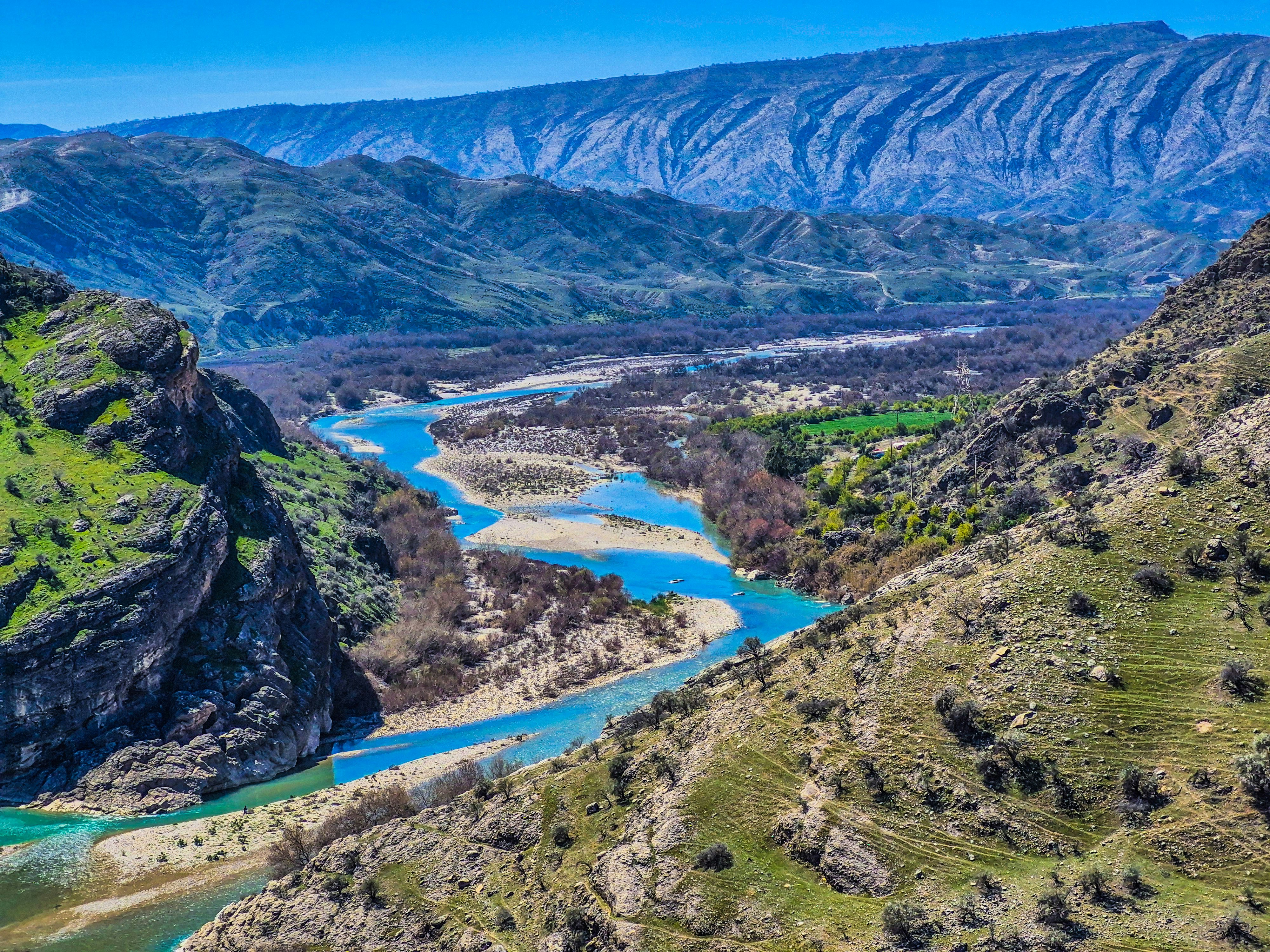 Winding turquoise river cutting through lush green valleys with distant mountains under a clear blue sky.