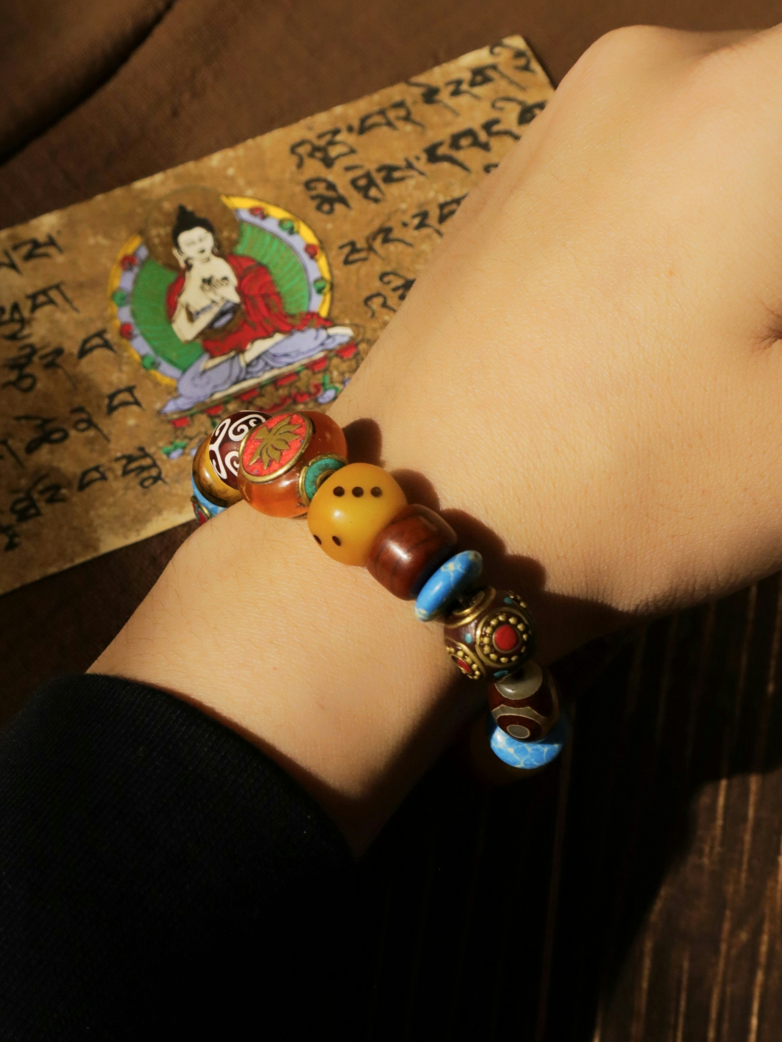 Colorful bead bracelet resting on a hand, with a decorative background featuring a Buddhist illustration and script.