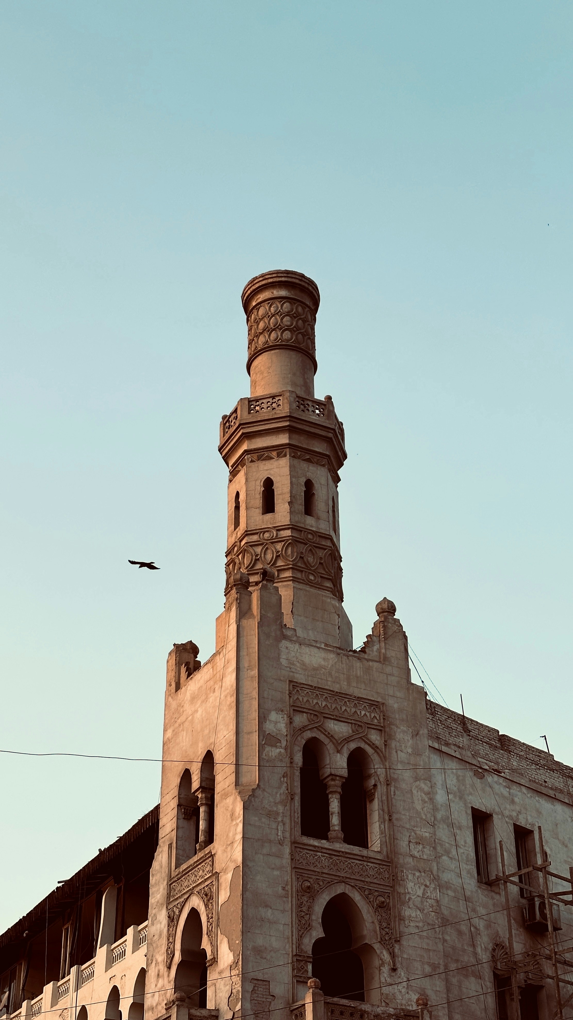 Intricate architectural tower rises against a clear sky, showcasing historical design elements. A lone bird flies nearby, adding a sense of life to the scene.