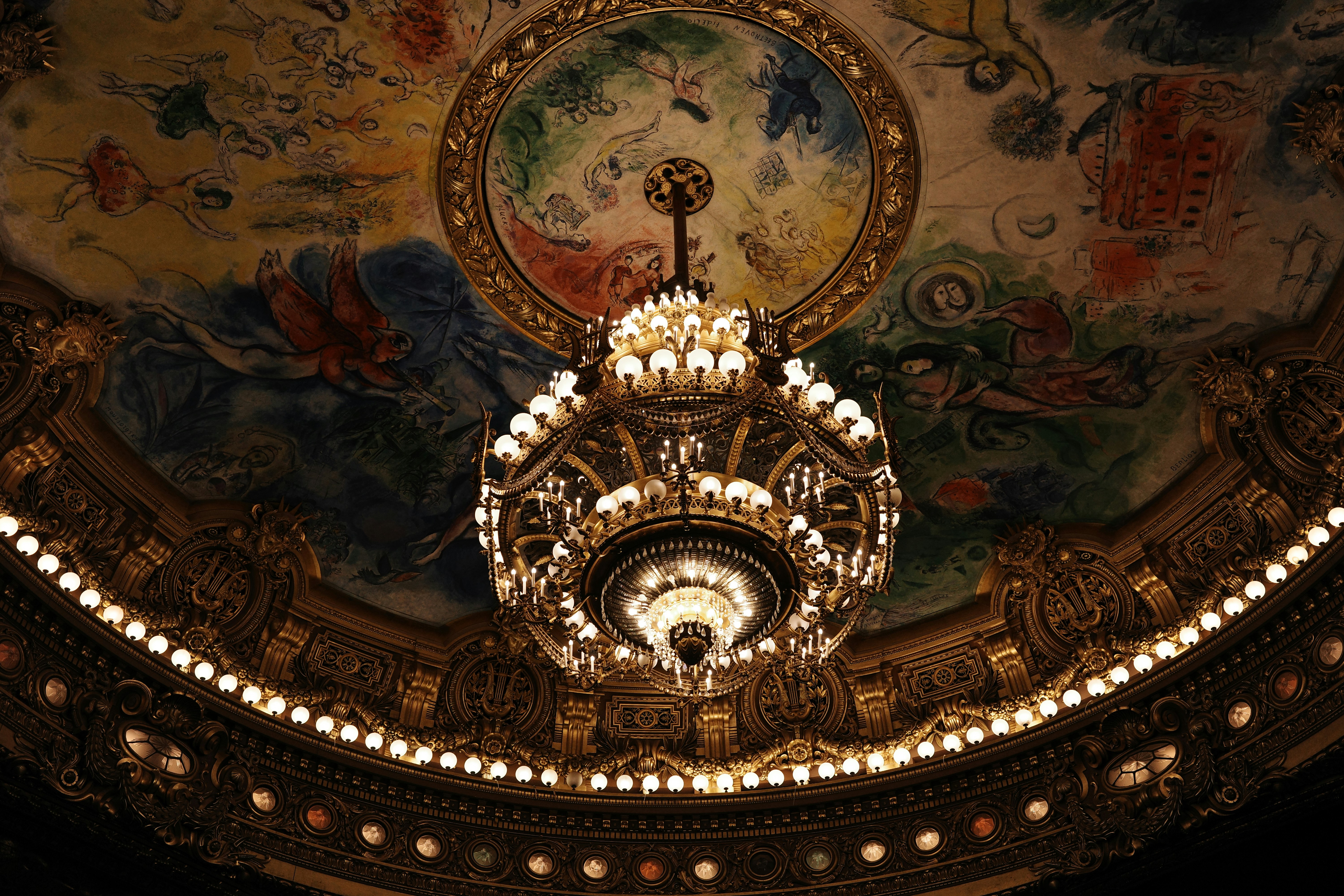 Ornate chandelier illuminates a vibrant, detailed ceiling mural with gold accents and rich colors.