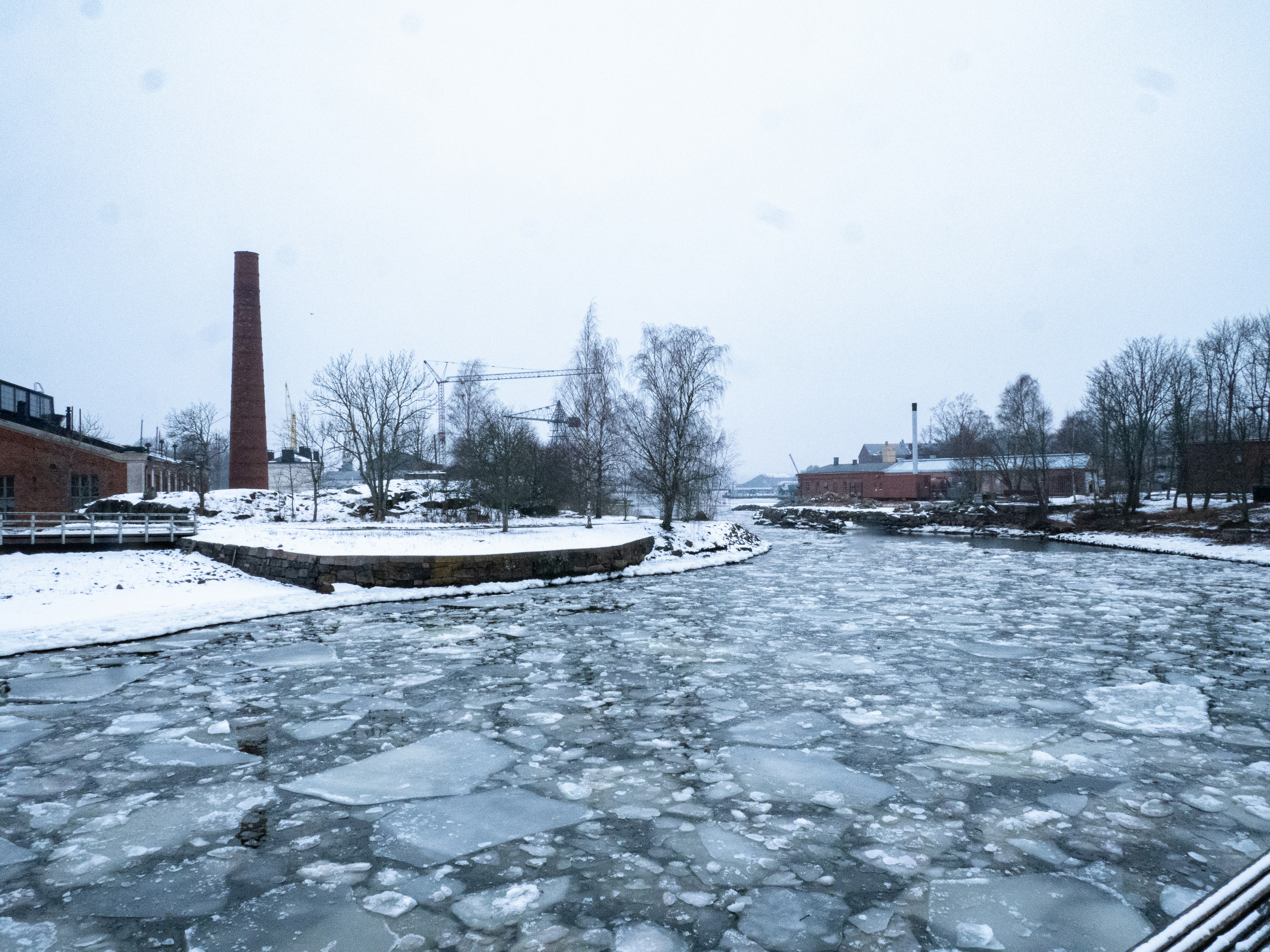 Frozen river flows past snow-covered trees and industrial buildings under a grey sky.