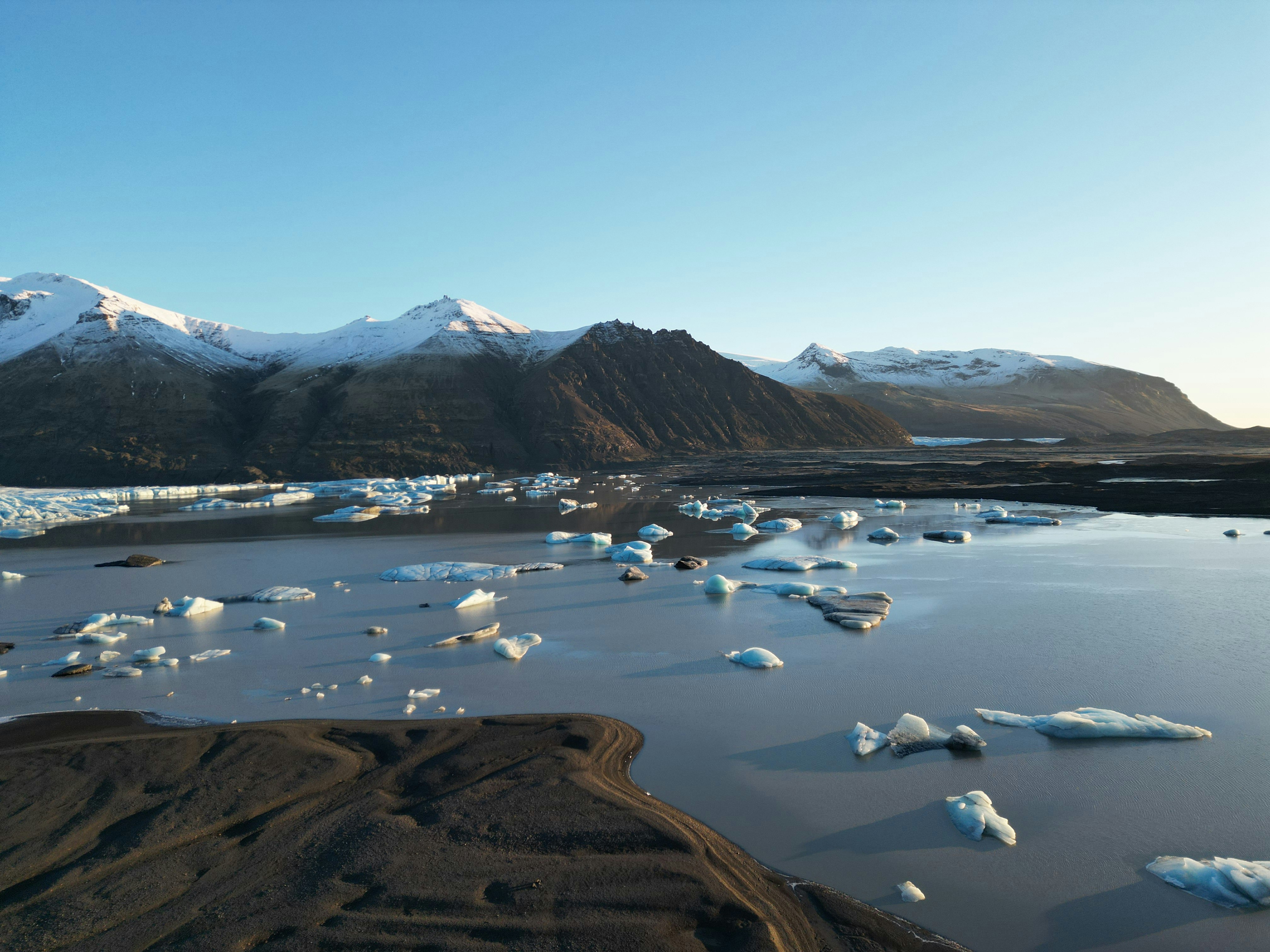 Icebergs float in a serene lake, with snow-capped mountains under a clear blue sky.