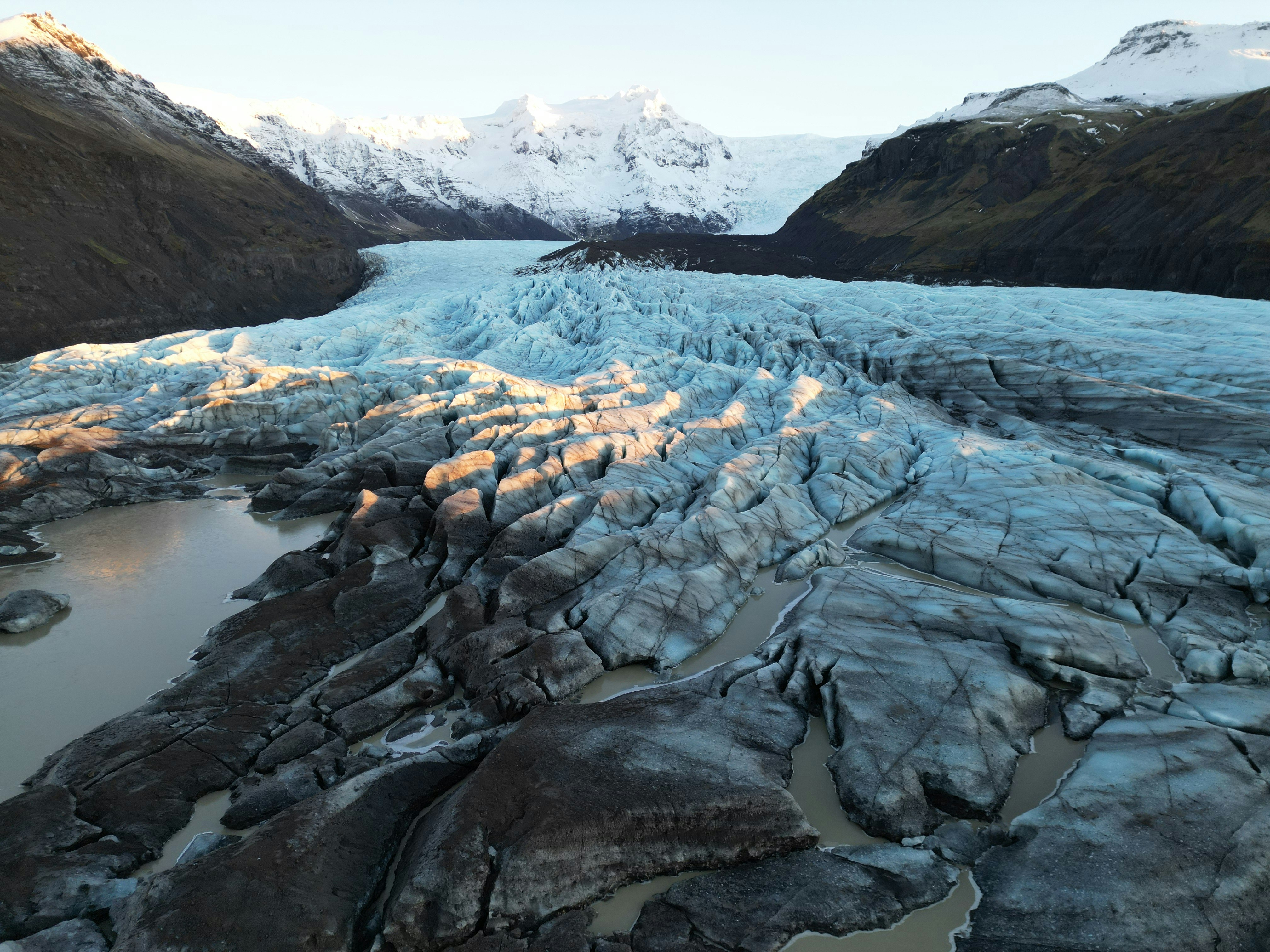 Expansive glacier with intricate patterns flows through rugged mountains under soft sunlight.