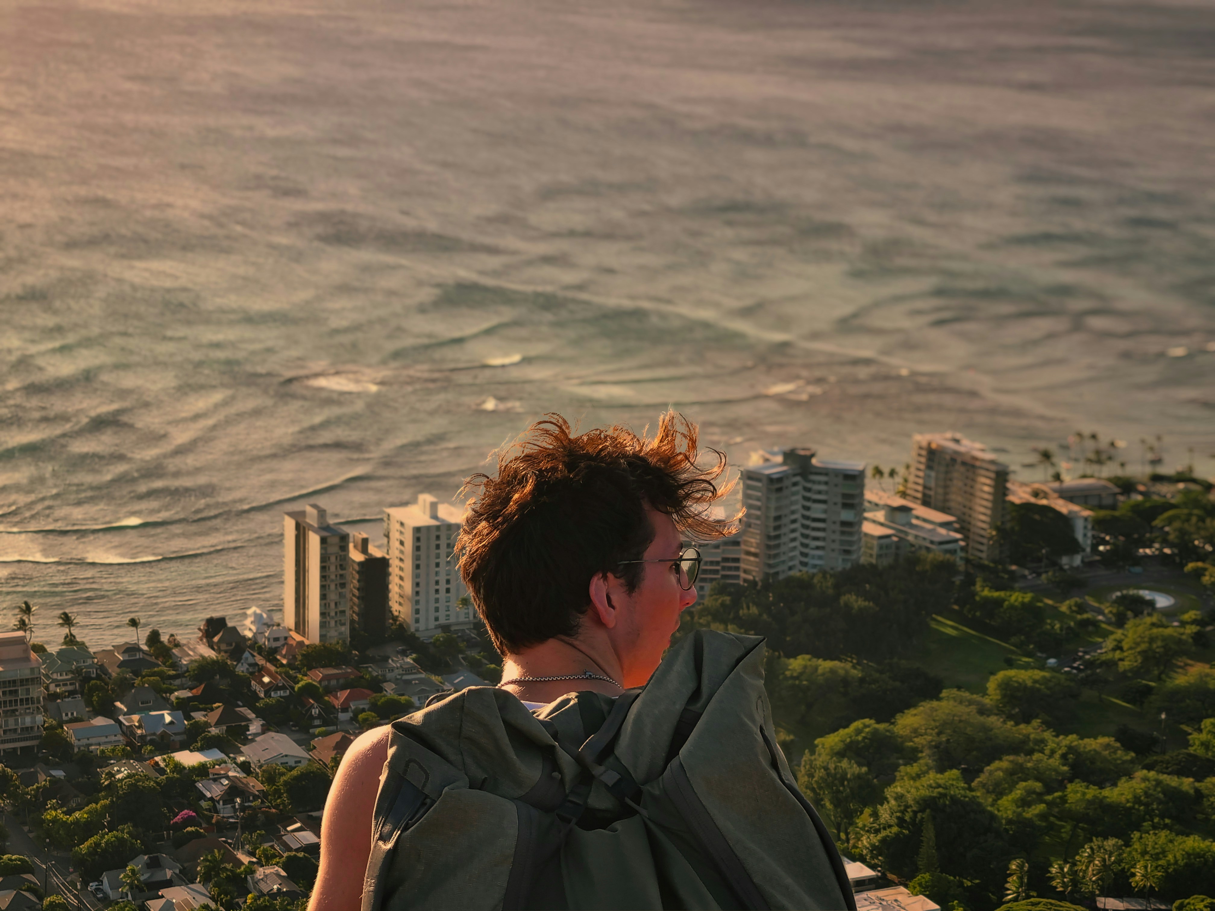 Person gazing at ocean waves and cityscape from a high vantage point during sunset.
