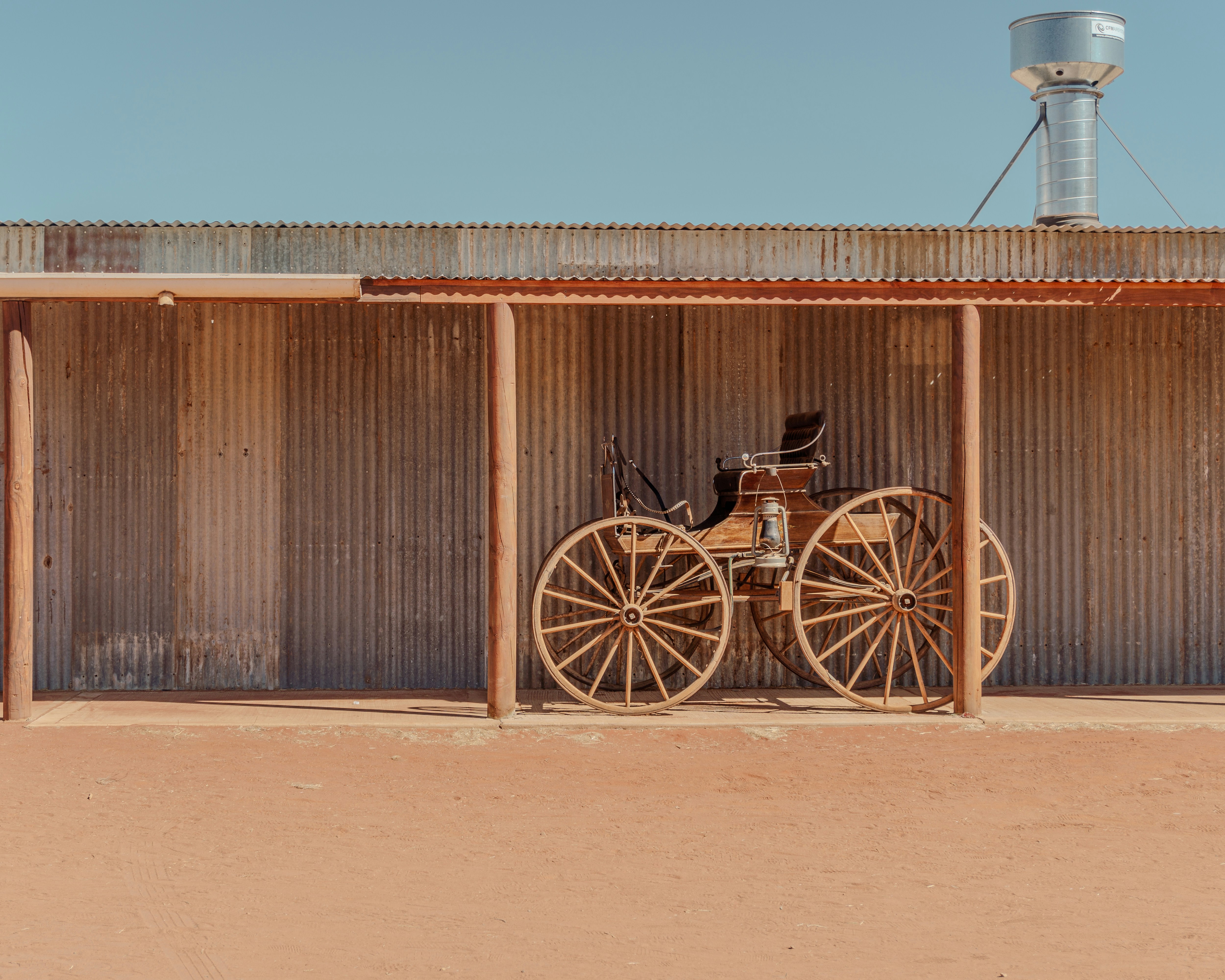 Vintage wooden carriage parked under a corrugated metal shed in a dusty landscape.