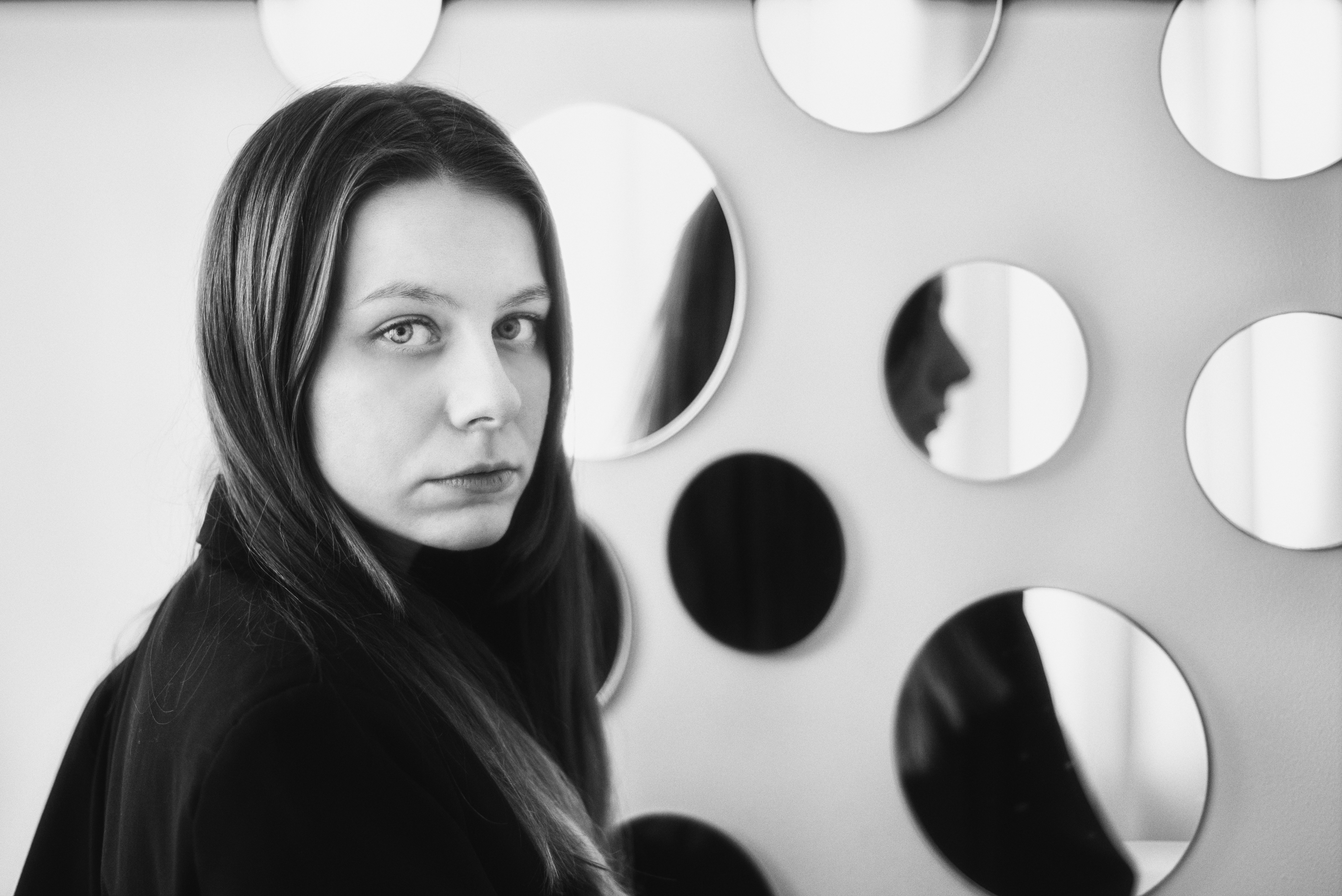 Woman with long hair stands beside a wall adorned with round mirrors, her reflection partially visible.
