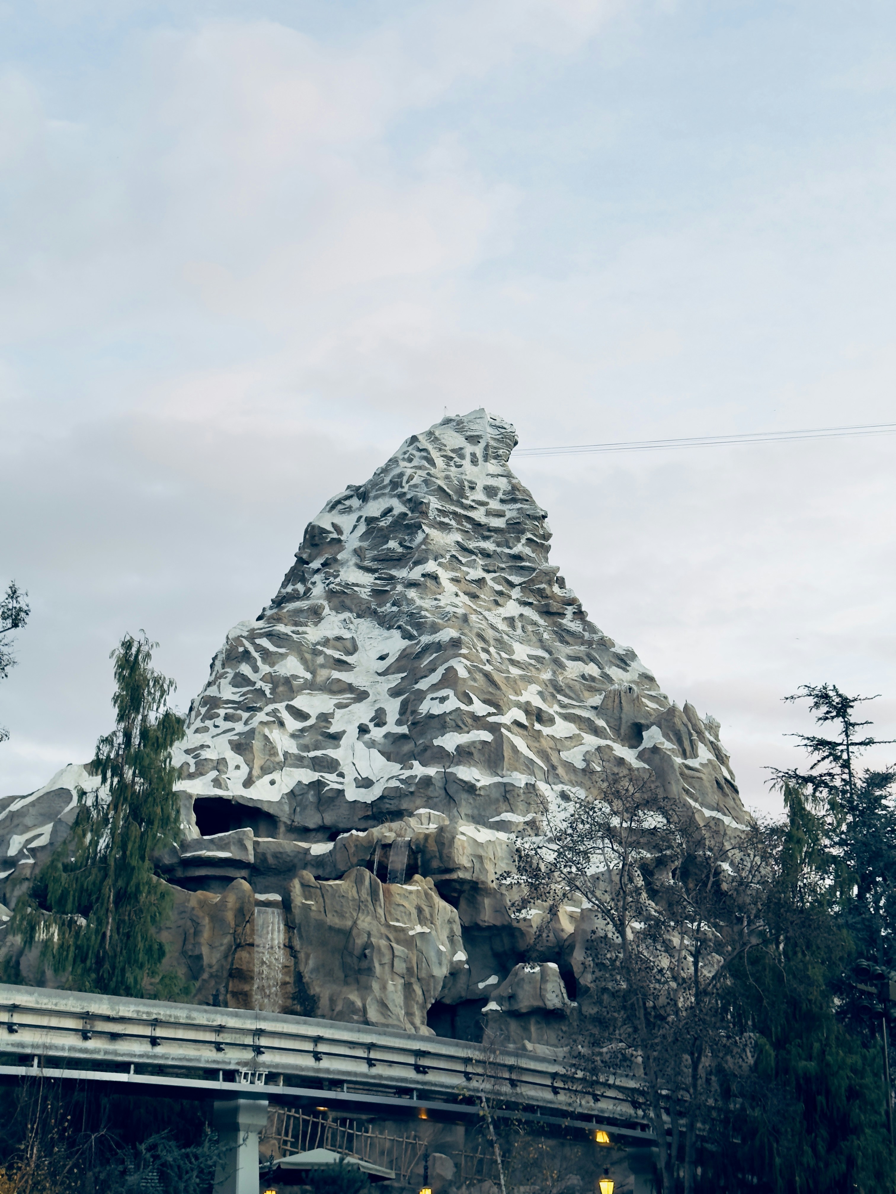 Photo captures a snow-dusted peak rising above a highway overpass, with trees in the foreground and a pale, cloudy sky.
