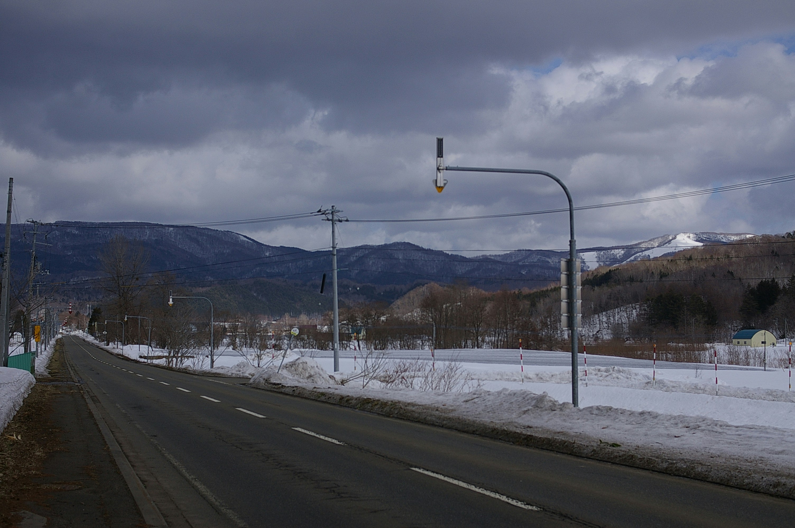 Snow-covered road leading towards distant mountains under a dramatic sky. Utility poles line the roadside, hinting at a quiet rural setting.