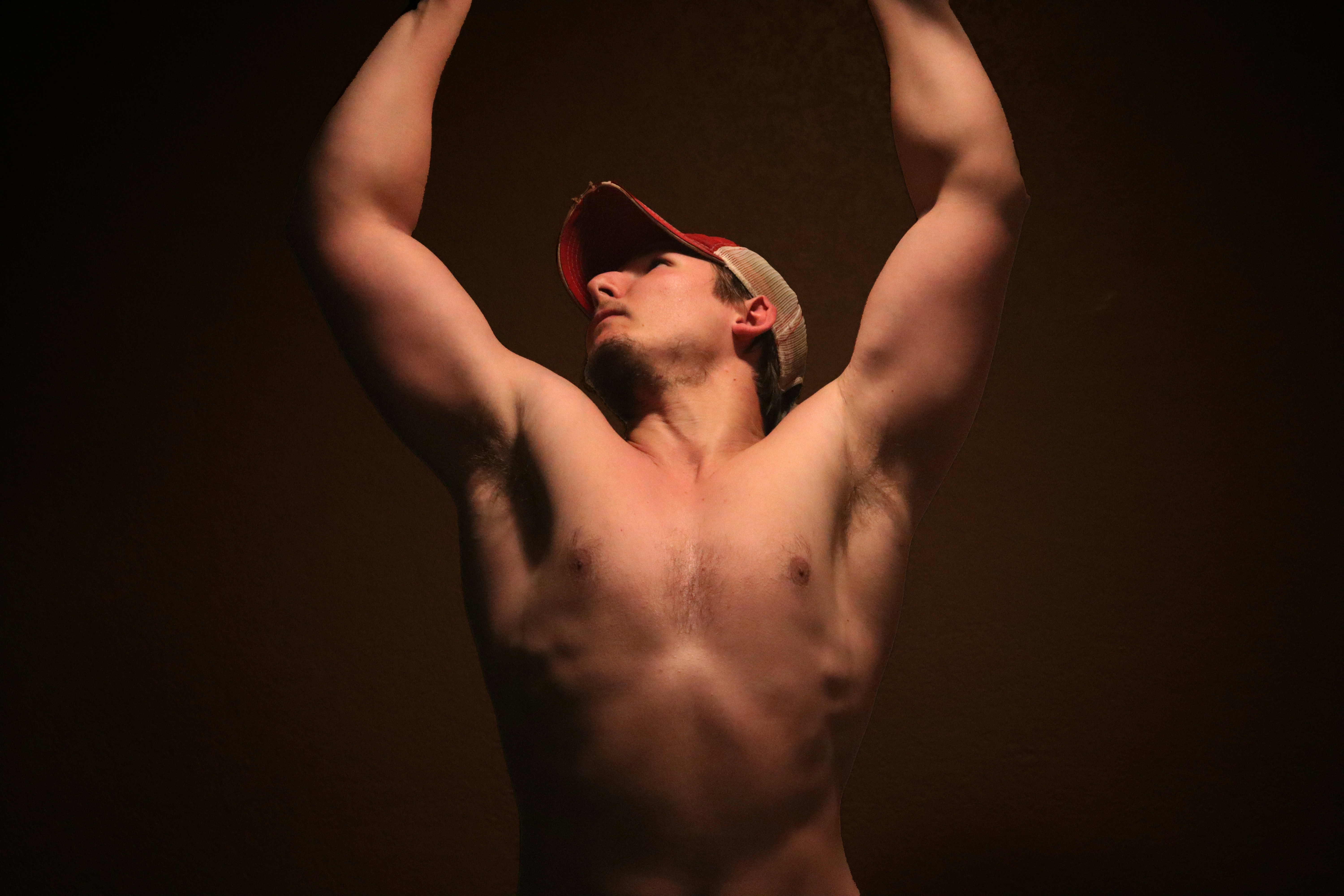 Shirtless man in a cap raising his arms against a dark background.
