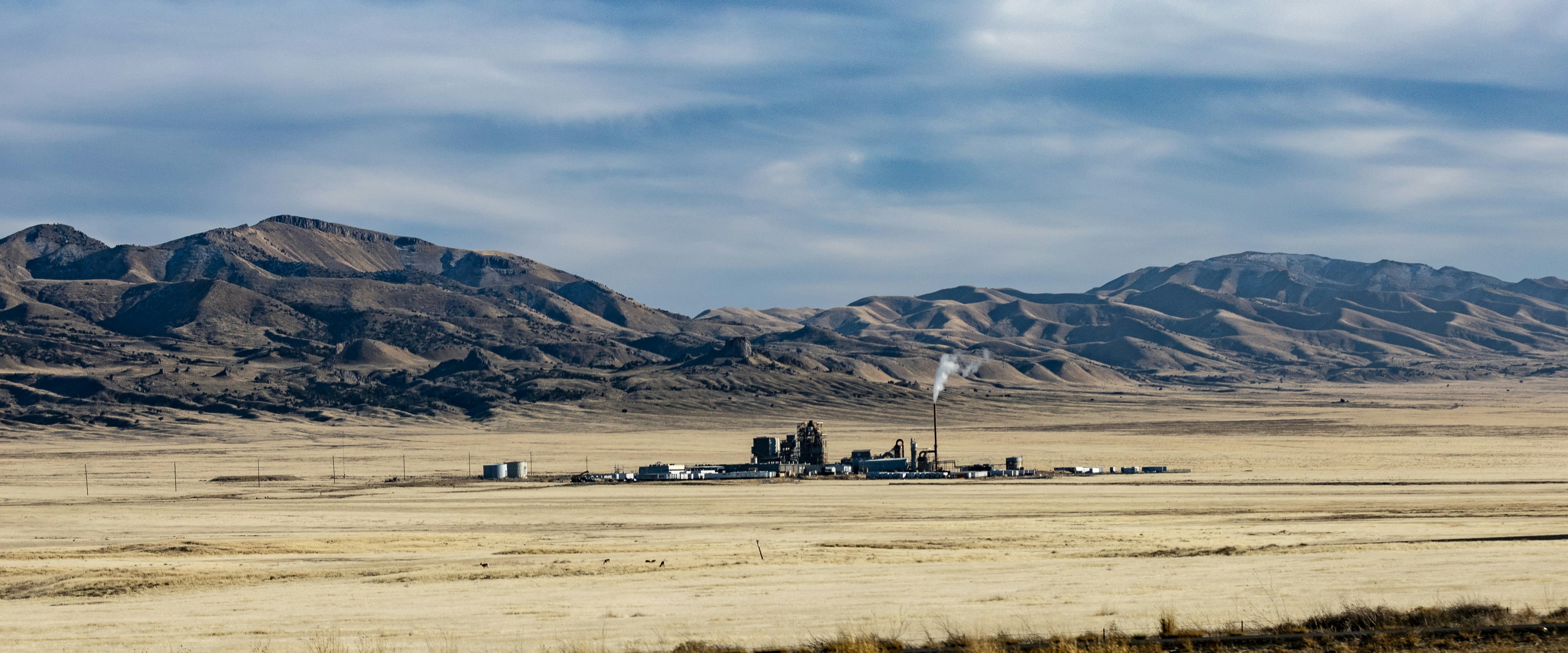 Remote refinery complex nestled in vast, dry fields with mountain backdrop under a cloudy sky.