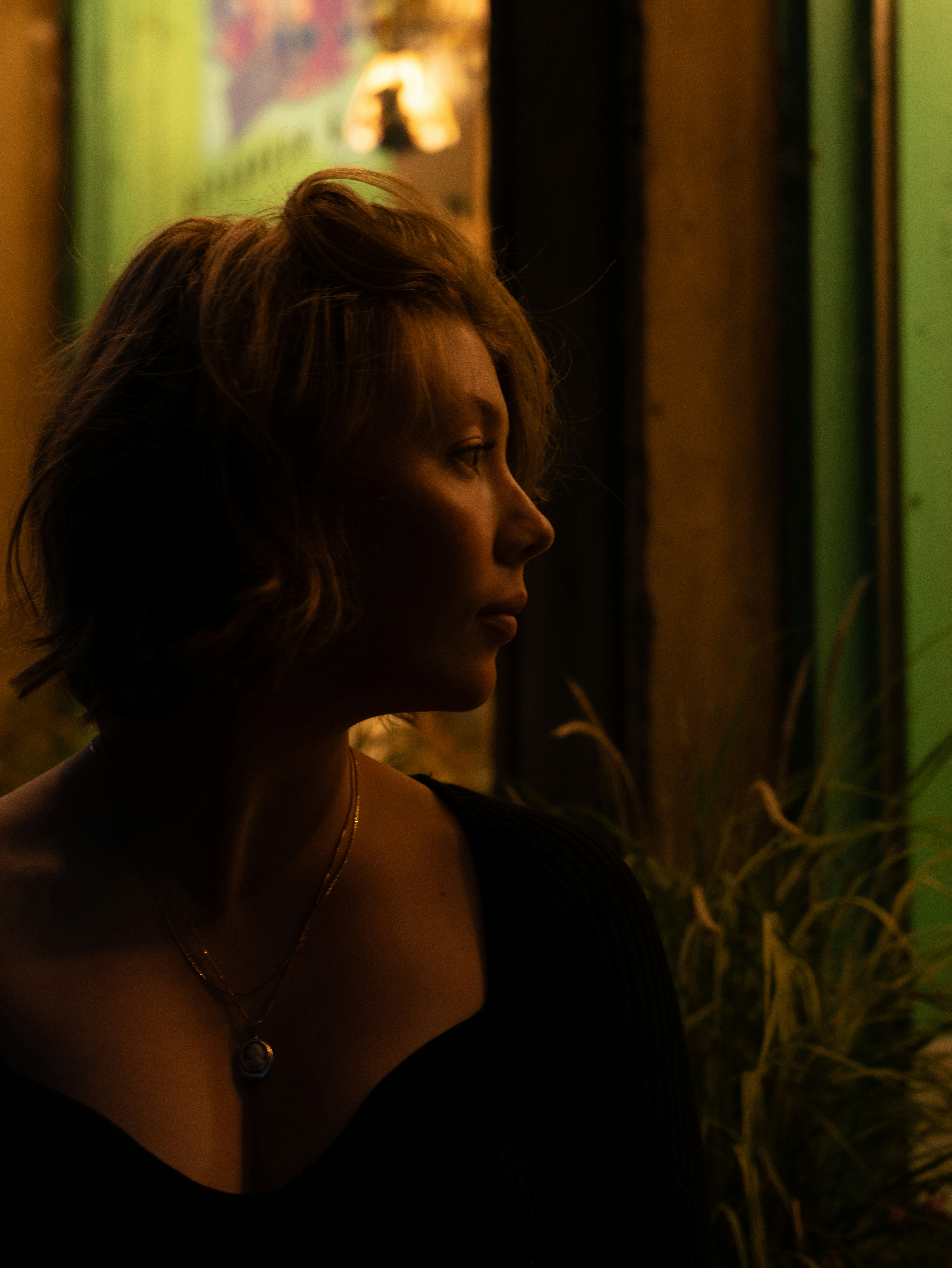 Woman gazing sideways in dim urban lighting near green and yellow walls.