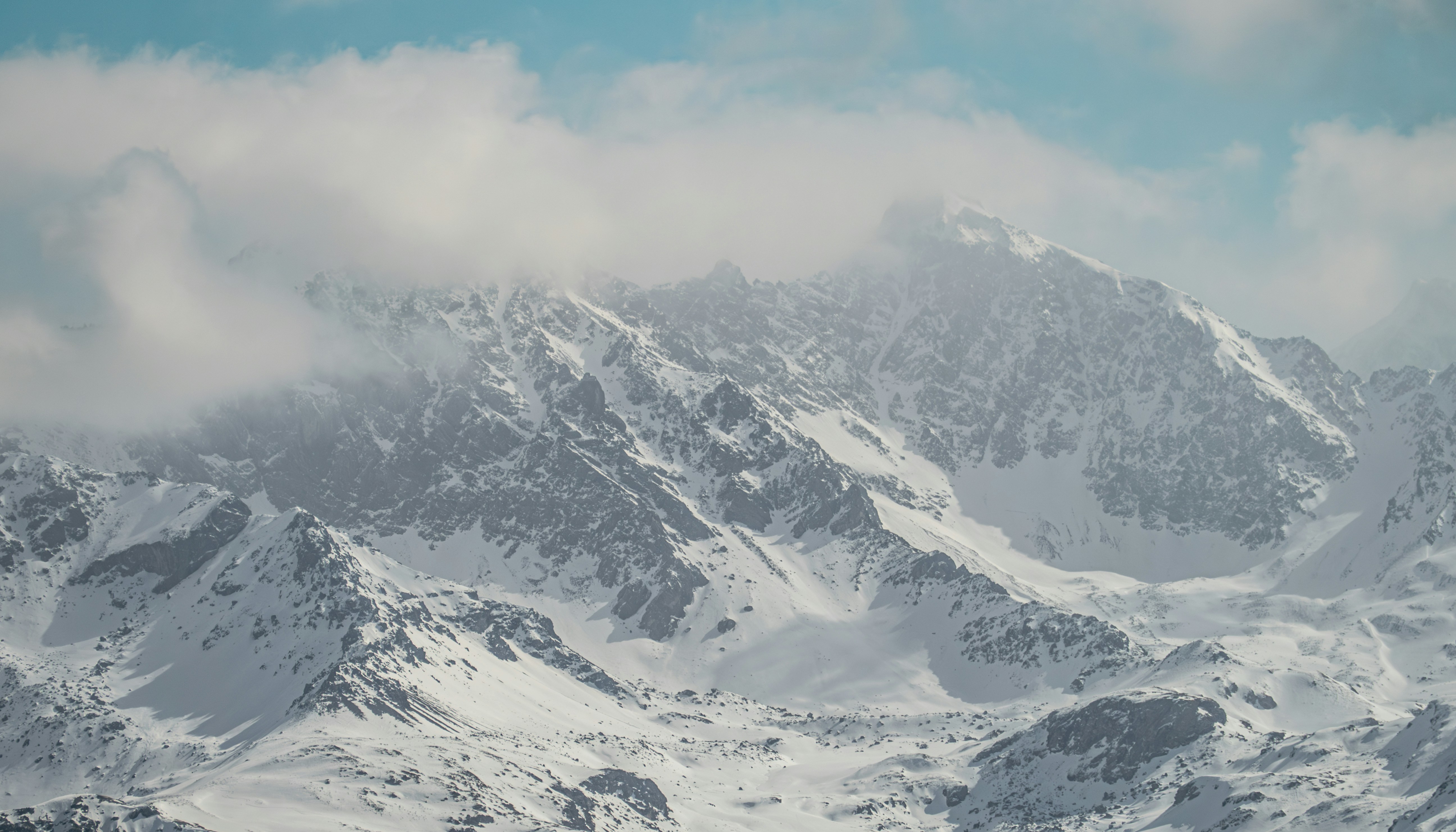 Snow-covered mountain range partially veiled by drifting clouds under a clear blue sky.