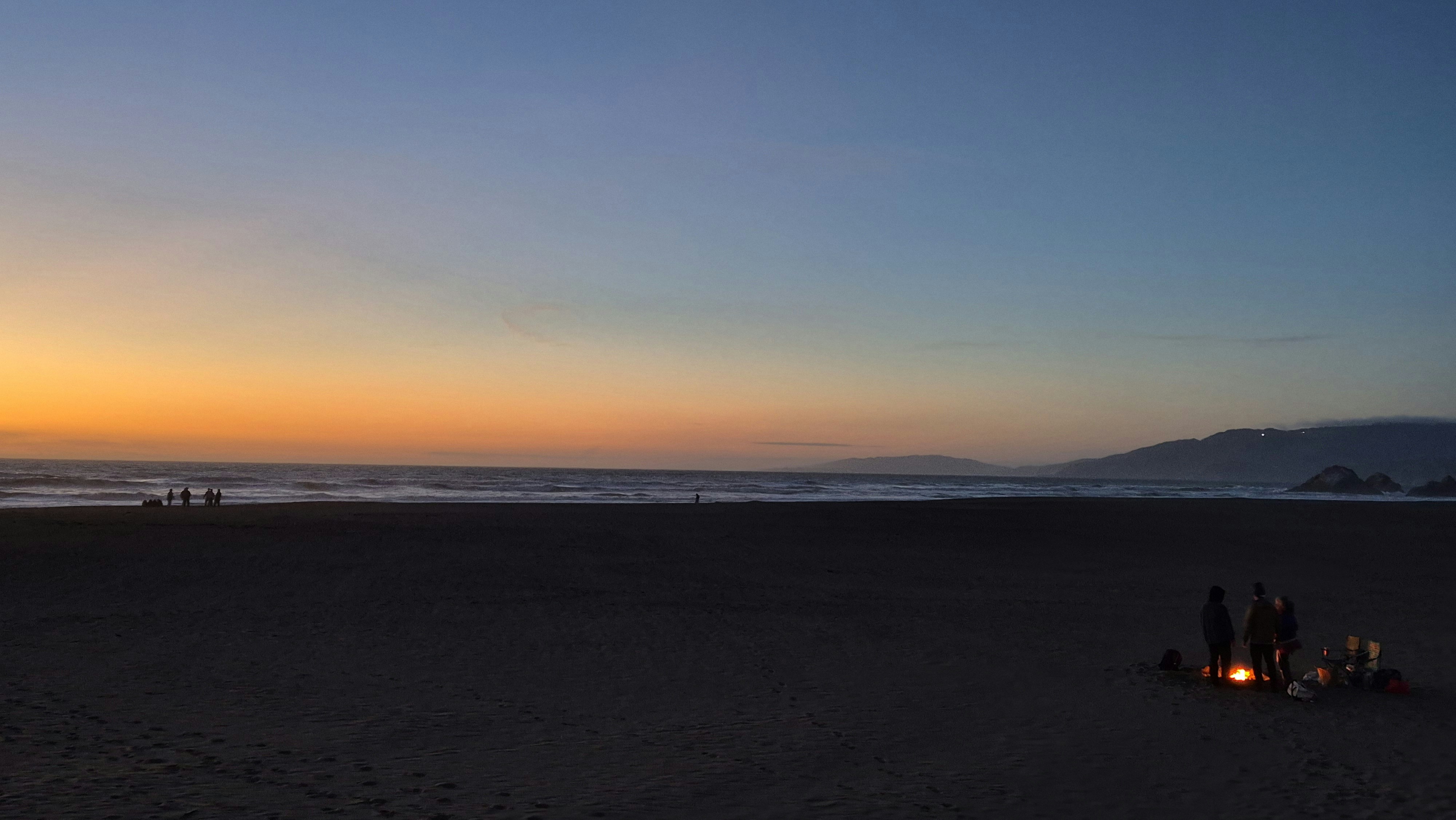 Beach at sunset with people around a small fire, silhouetted against the vibrant sky.