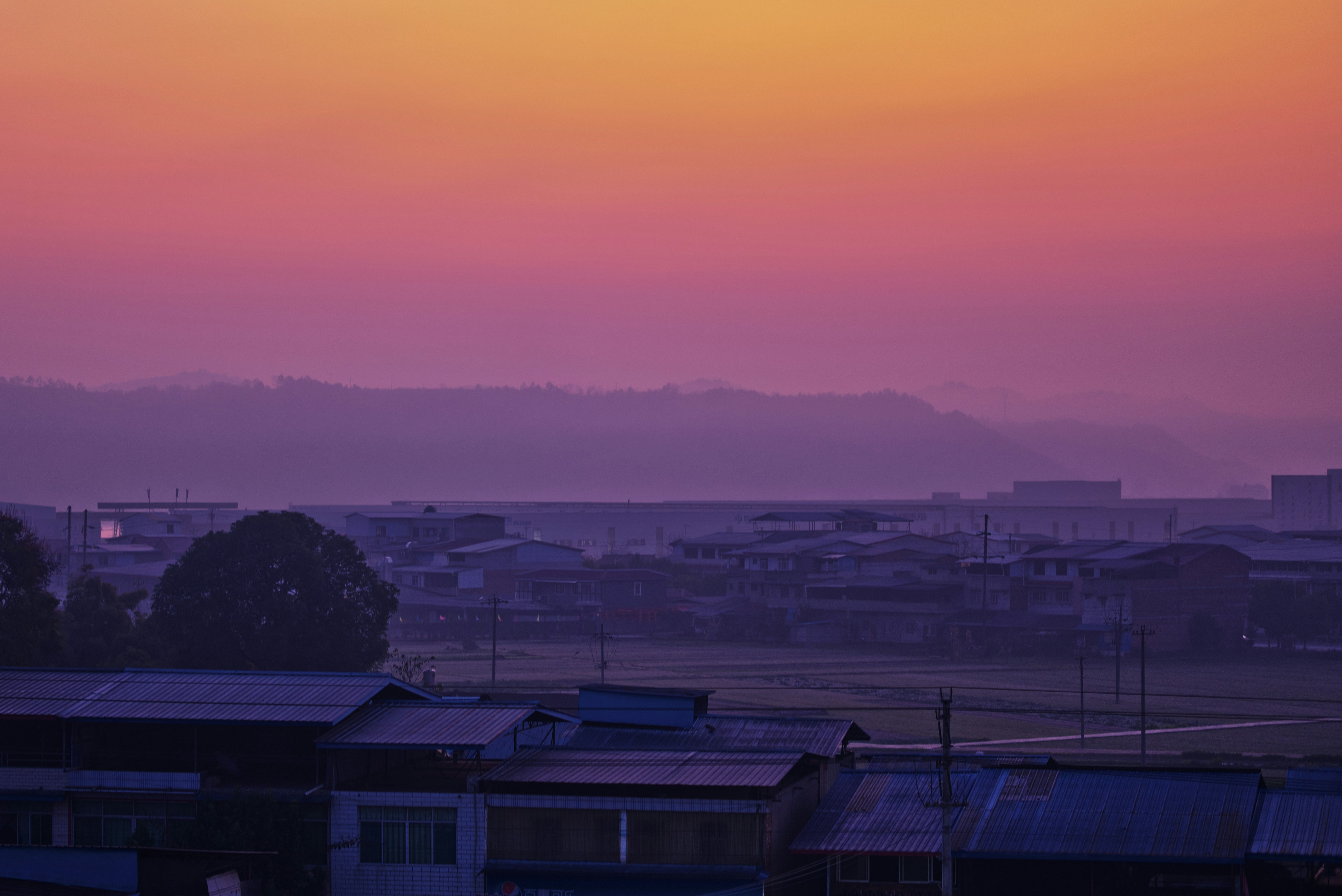 Vibrant sunset sky casting a warm glow over silhouetted rooftops and distant hills.