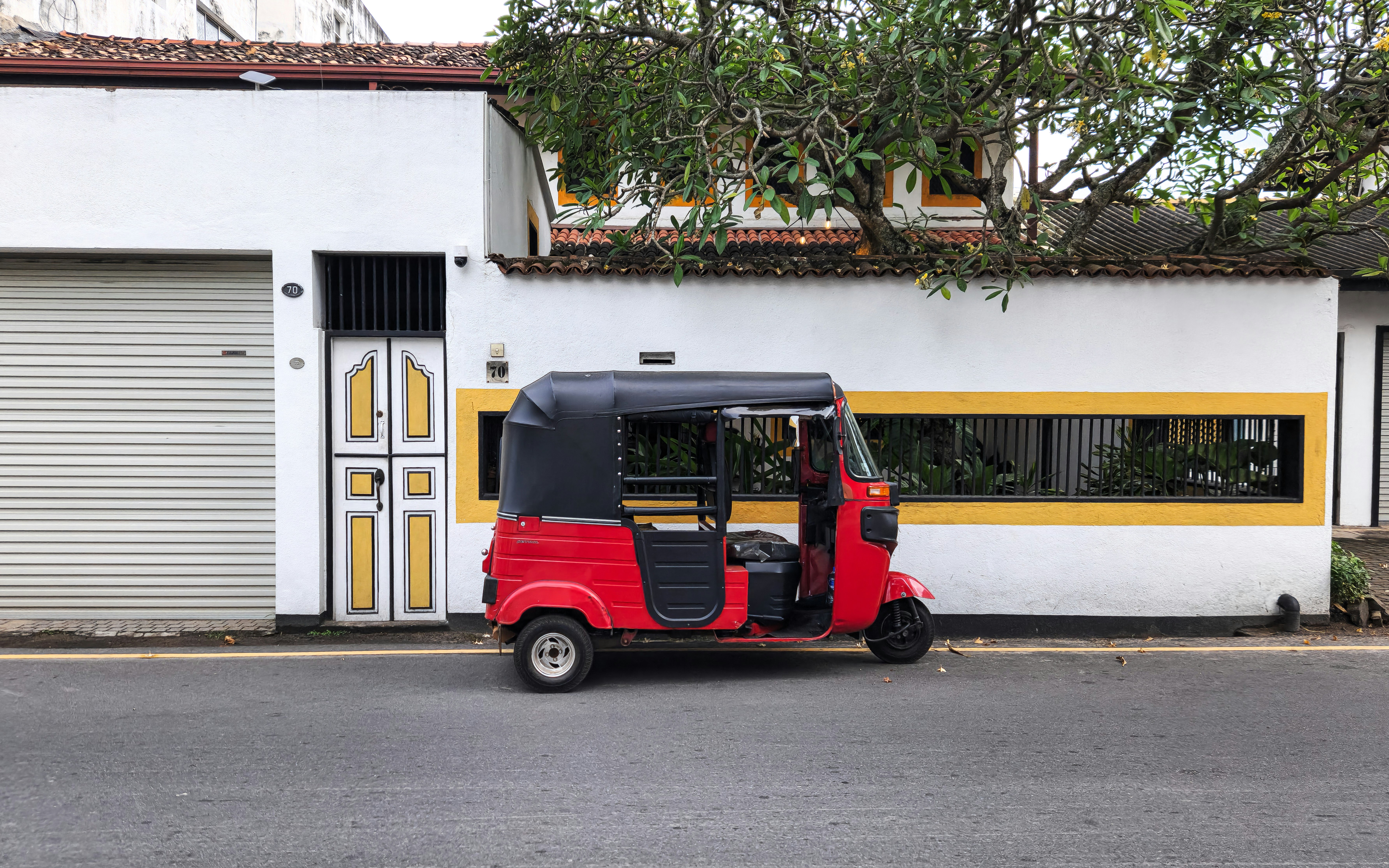 Red tuk-tuk parked against a white wall with yellow accents in Colombo, under a leafy tree.