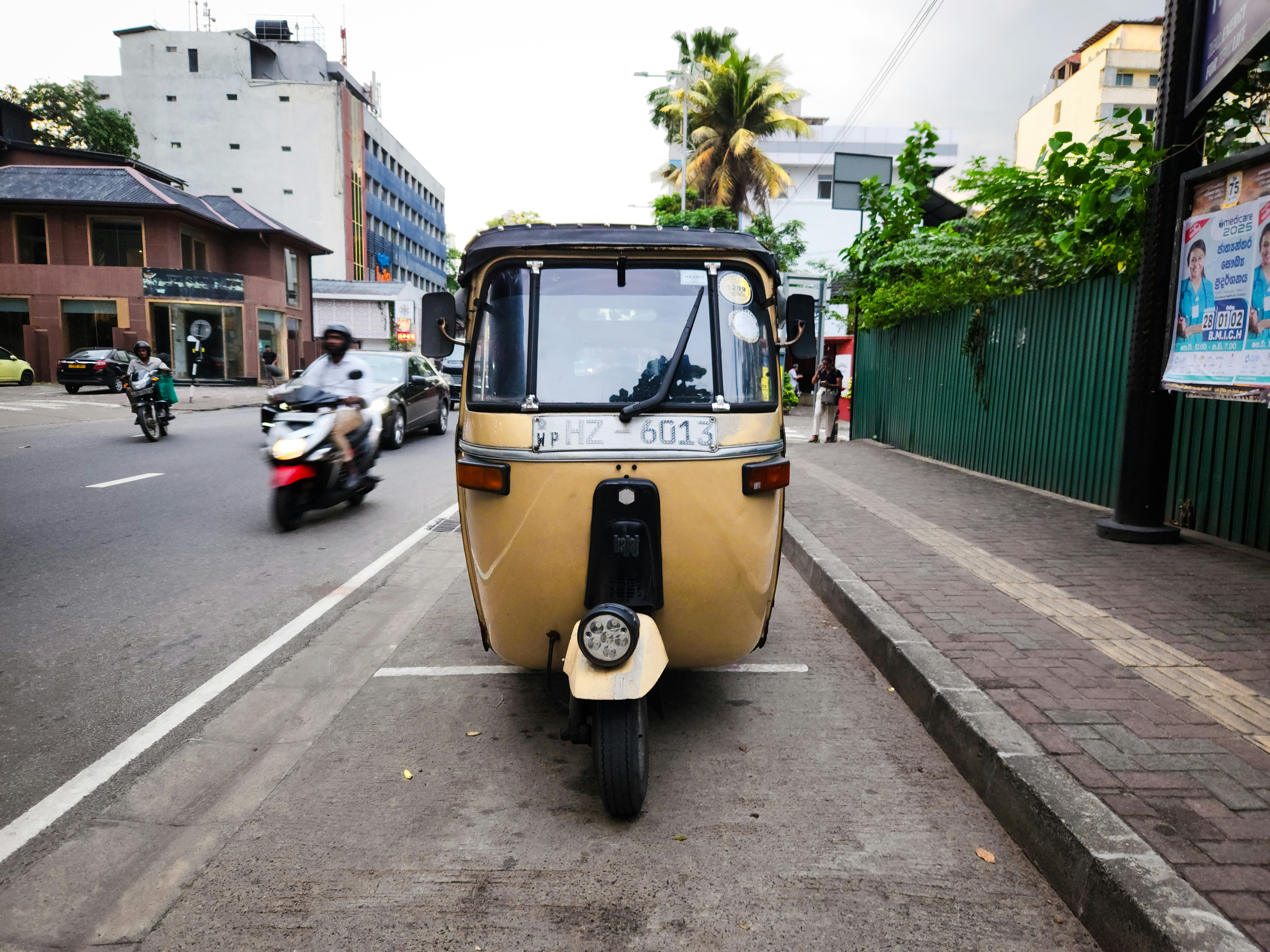Front view of a beige tuk-tuk parked on an urban street with blurred motion of passing vehicles.