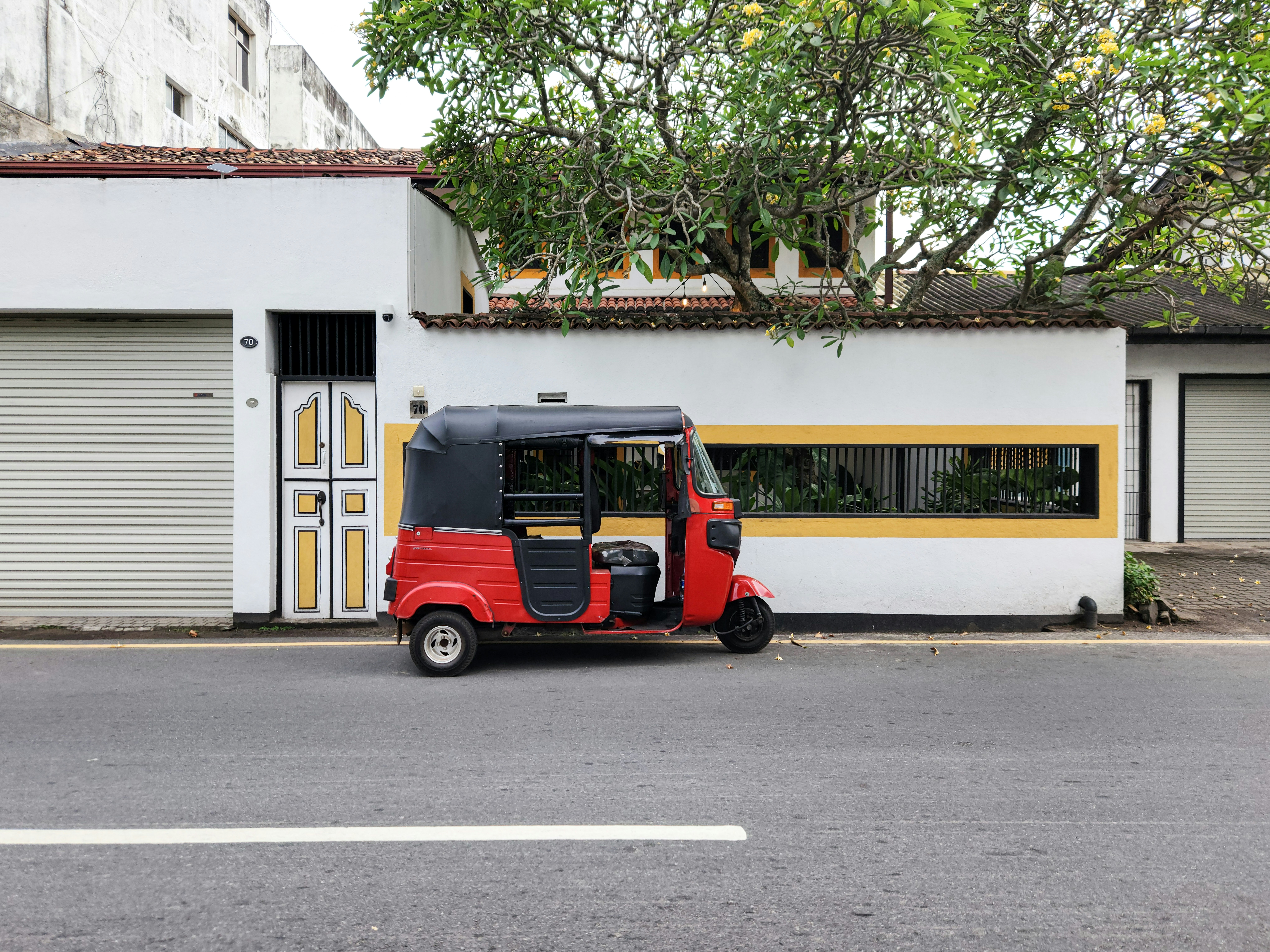 Red tuk-tuk parked against a white building with yellow accents and a leafy backdrop.