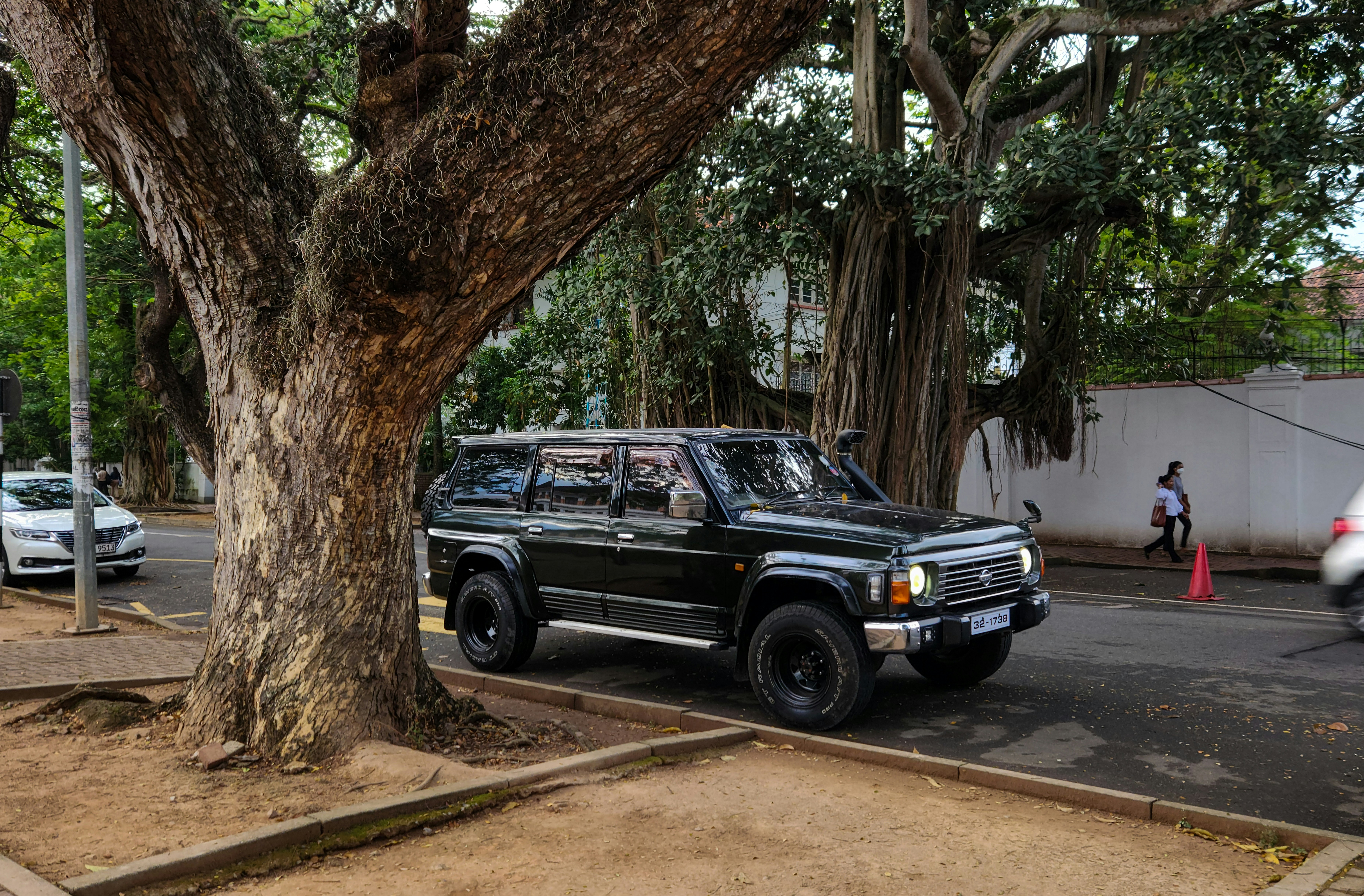 Black Nissan SUV parked under large trees with dense foliage on a paved street in Colombo.