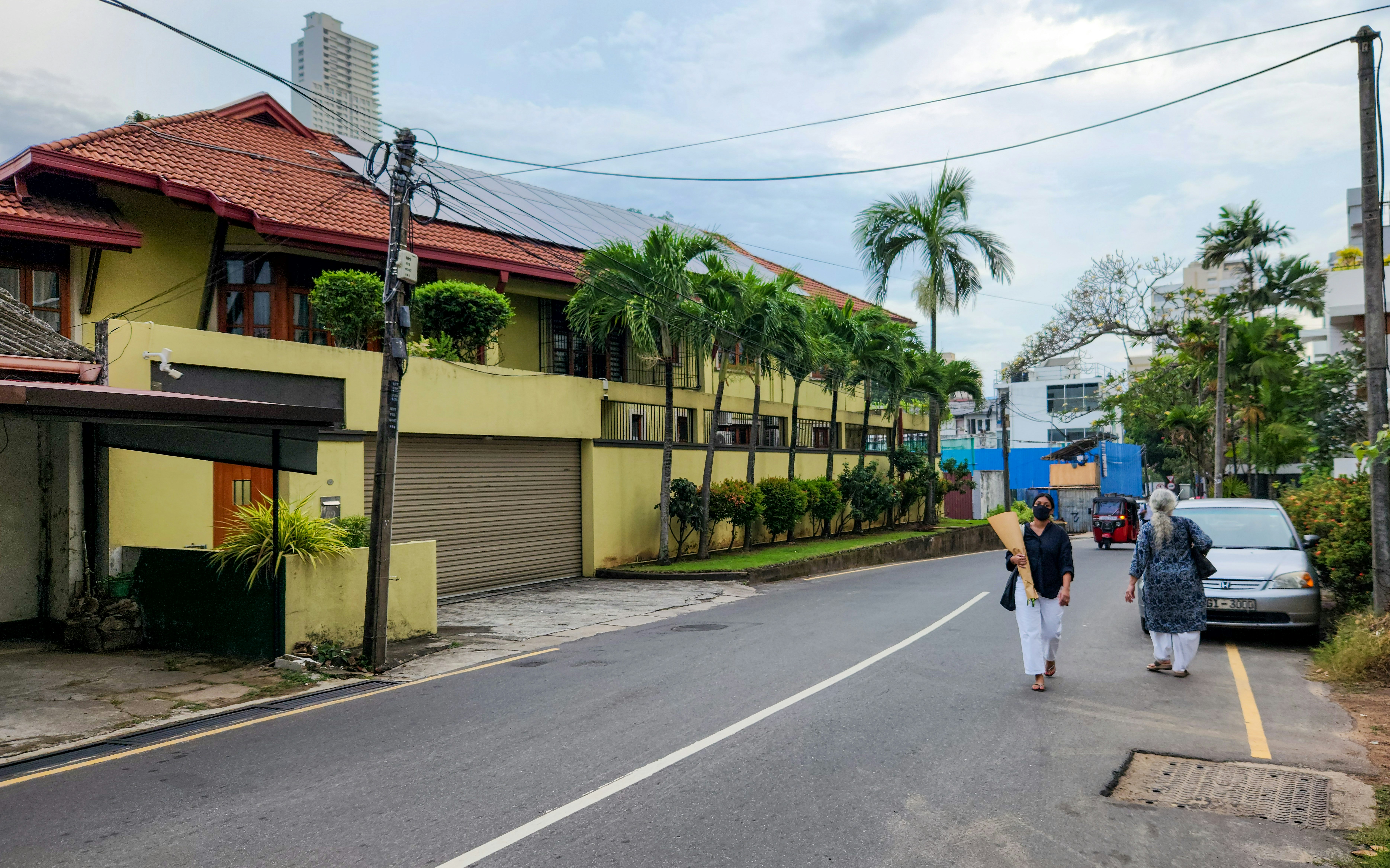 Two people walking along a tree-lined street beside yellow buildings under a cloudy sky.