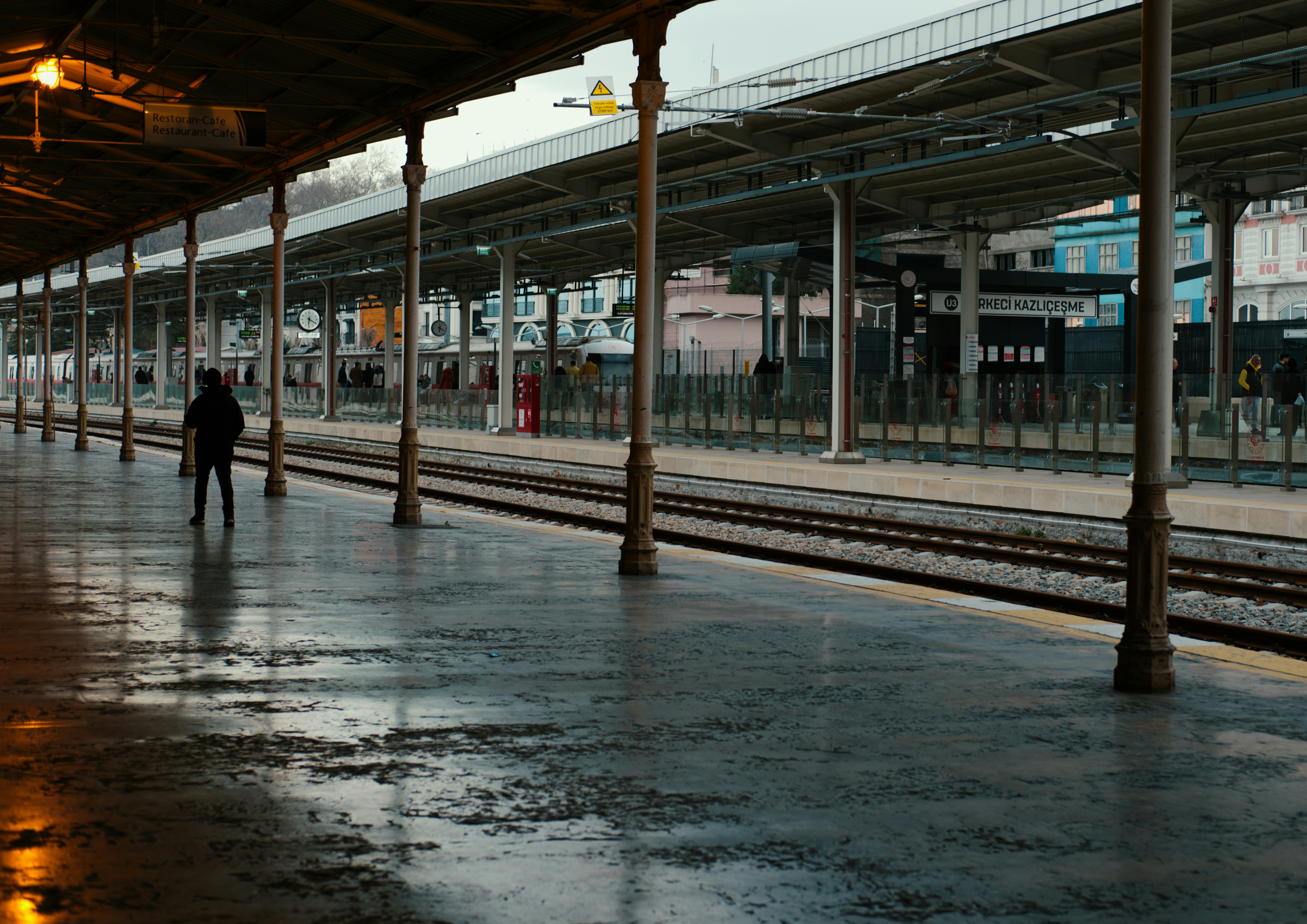 Silhouetted figure stands on a wet train platform under dim lighting, reflecting the quiet ambiance.