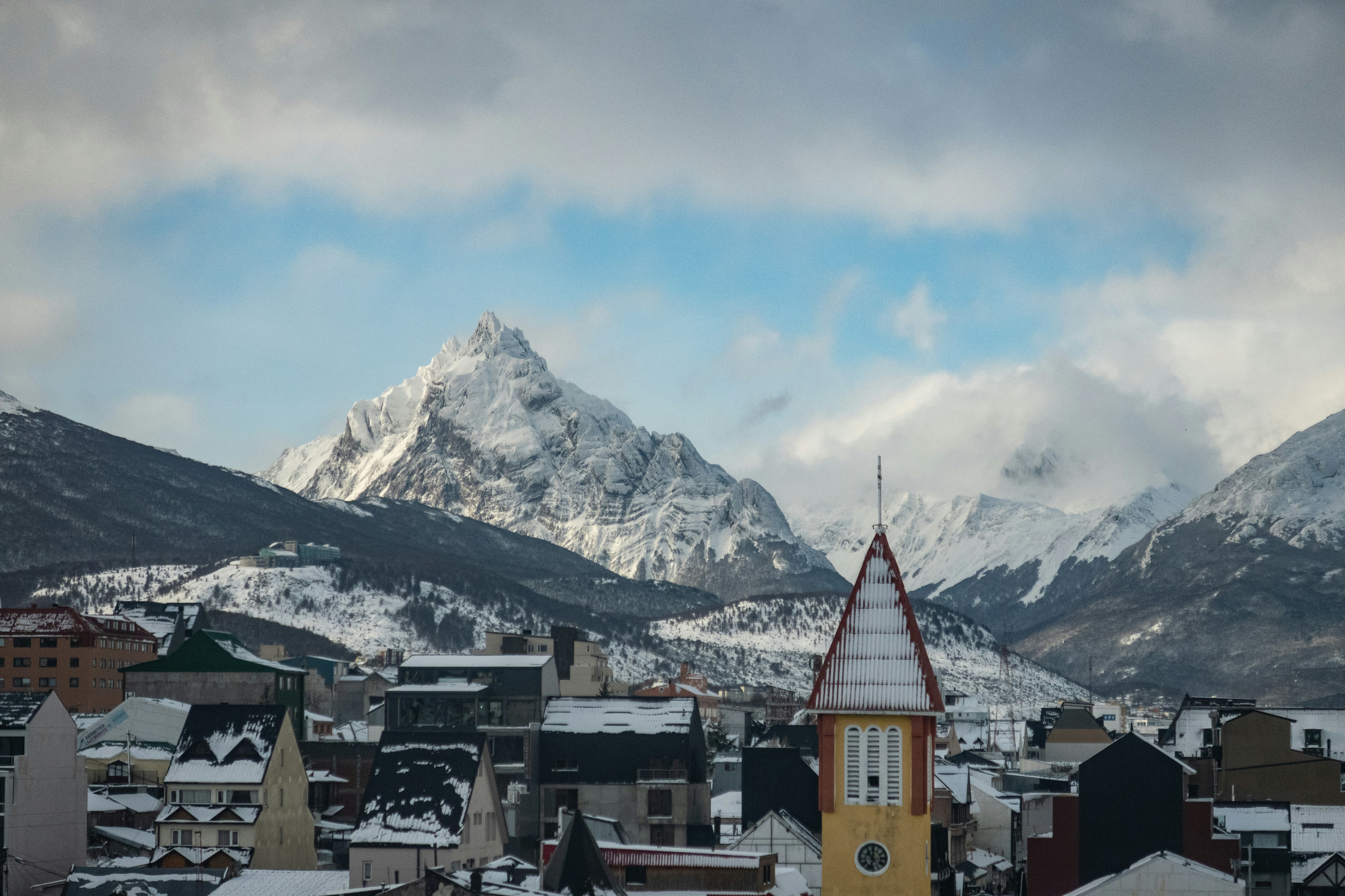 Snow-covered mountain towering over Ushuaia's vibrant rooftops under a partly cloudy sky.