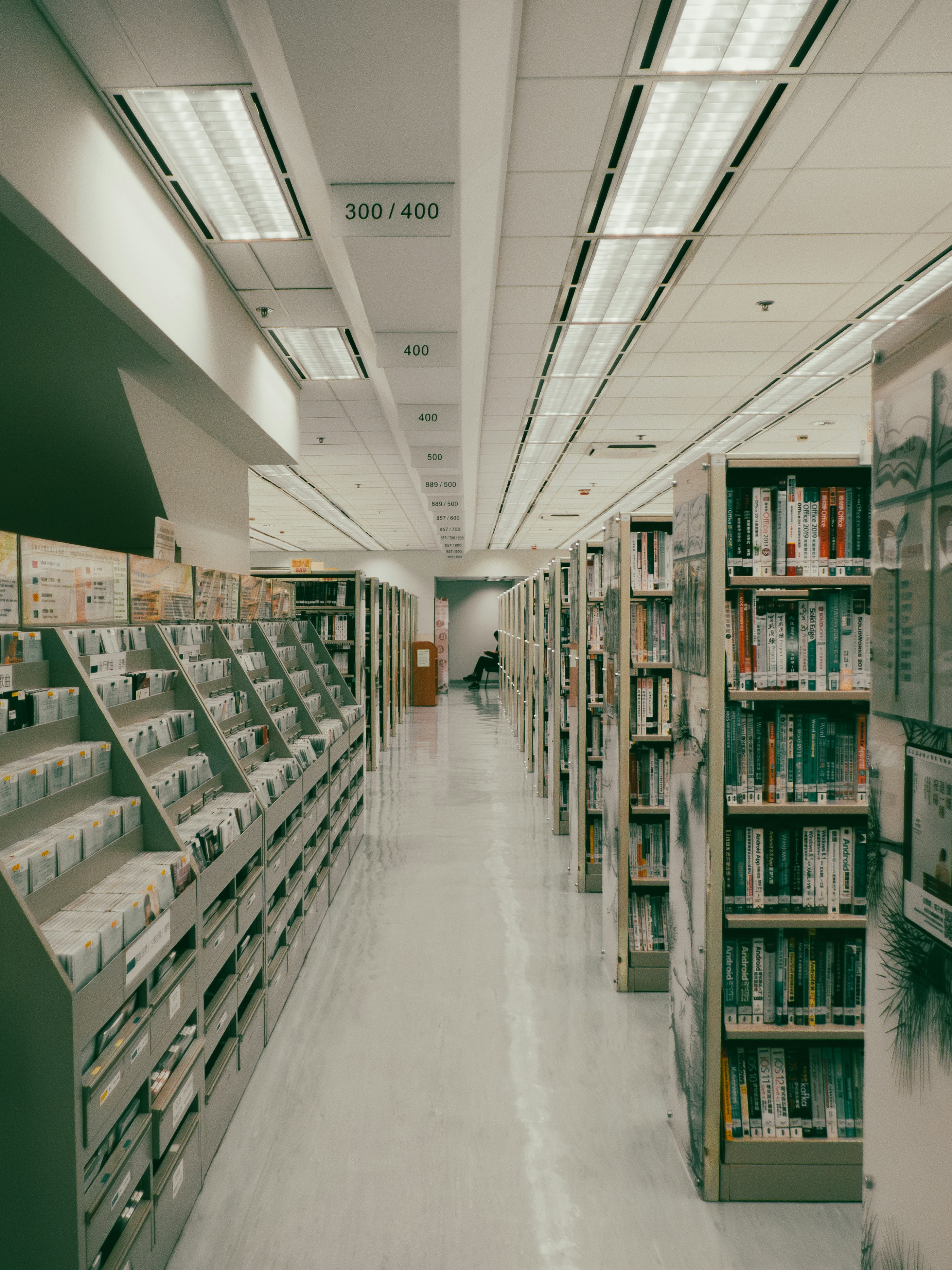 Wide-shot photograph of a modern library aisle with long shelves and bright ceiling lights, receding toward a distant figure at the end.