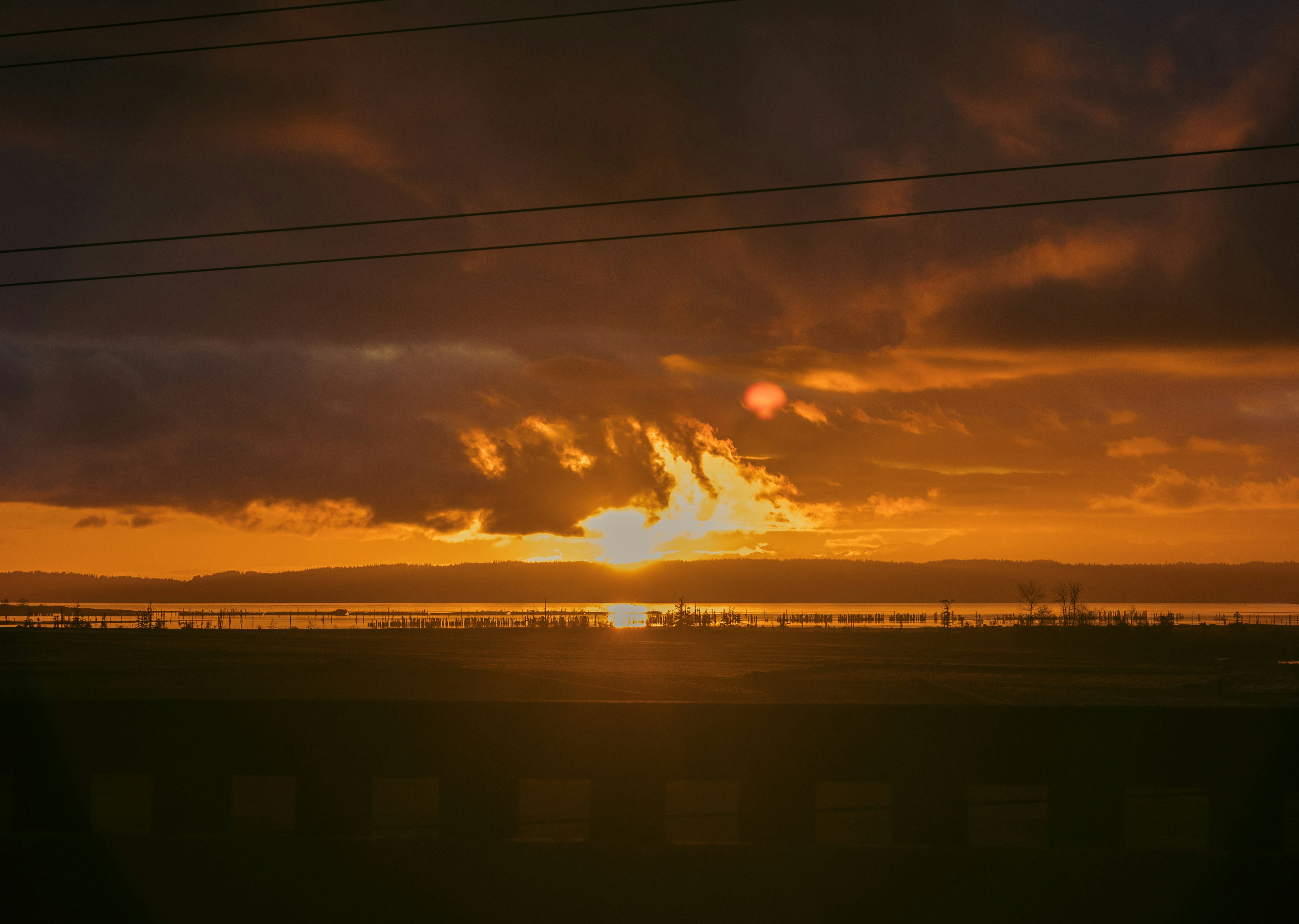Orange sunset over a calm body of water with silhouetted shoreline and power lines.