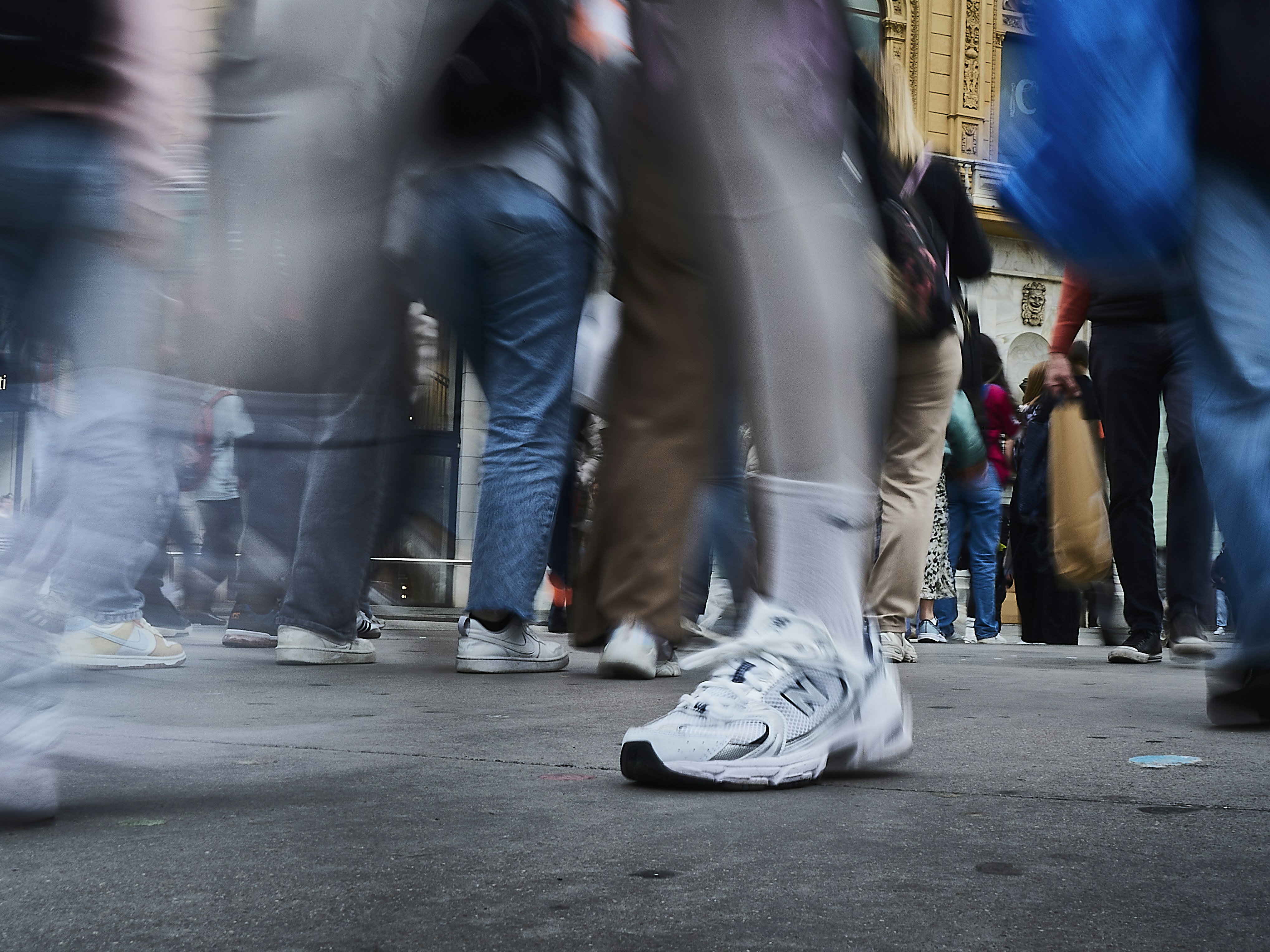 Blurred legs of pedestrians in motion on a busy city street with ornate building facade in the background.