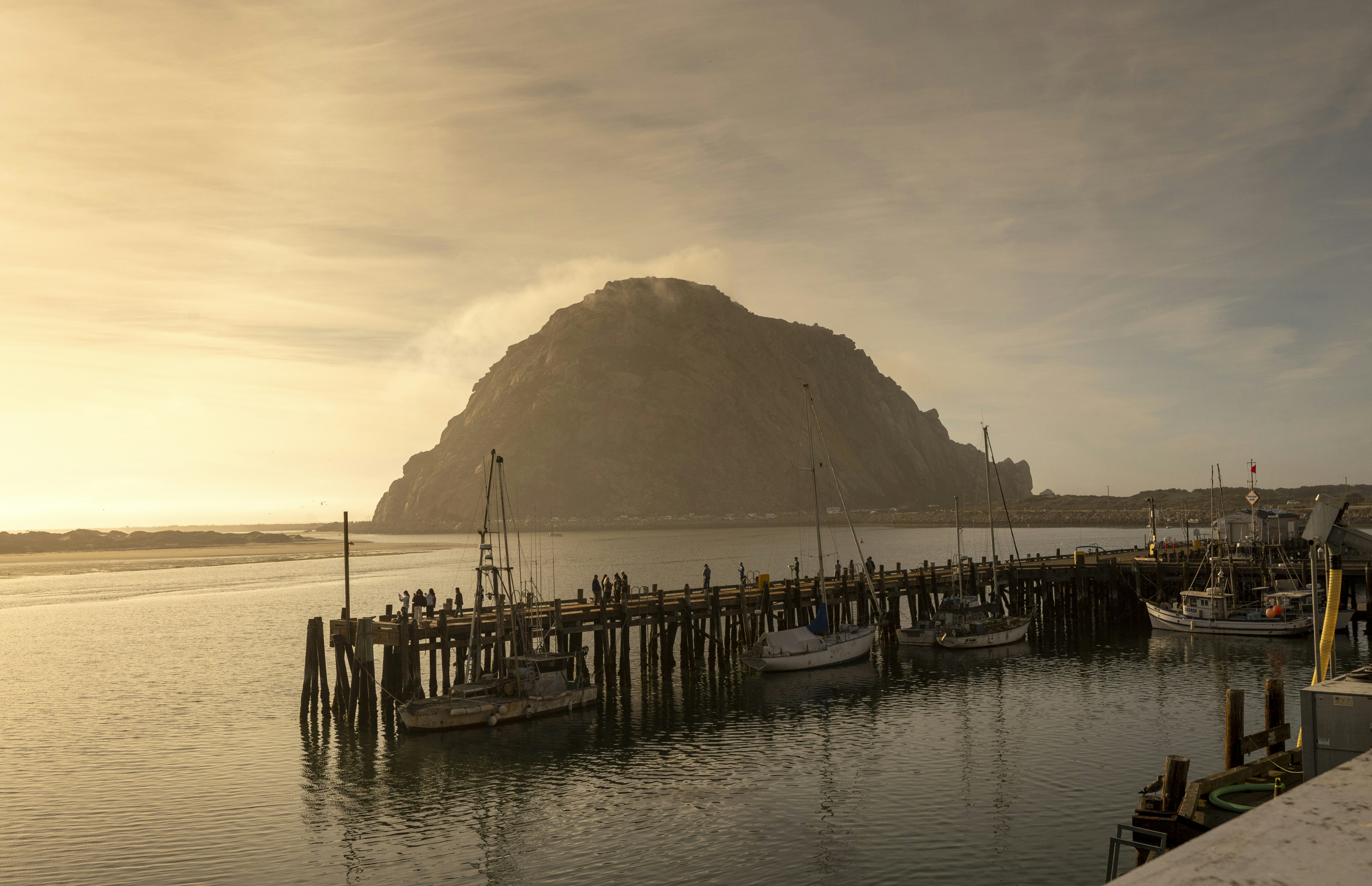 Fishing boats moored at a wooden pier with a towering rock formation in the background under a golden sky.