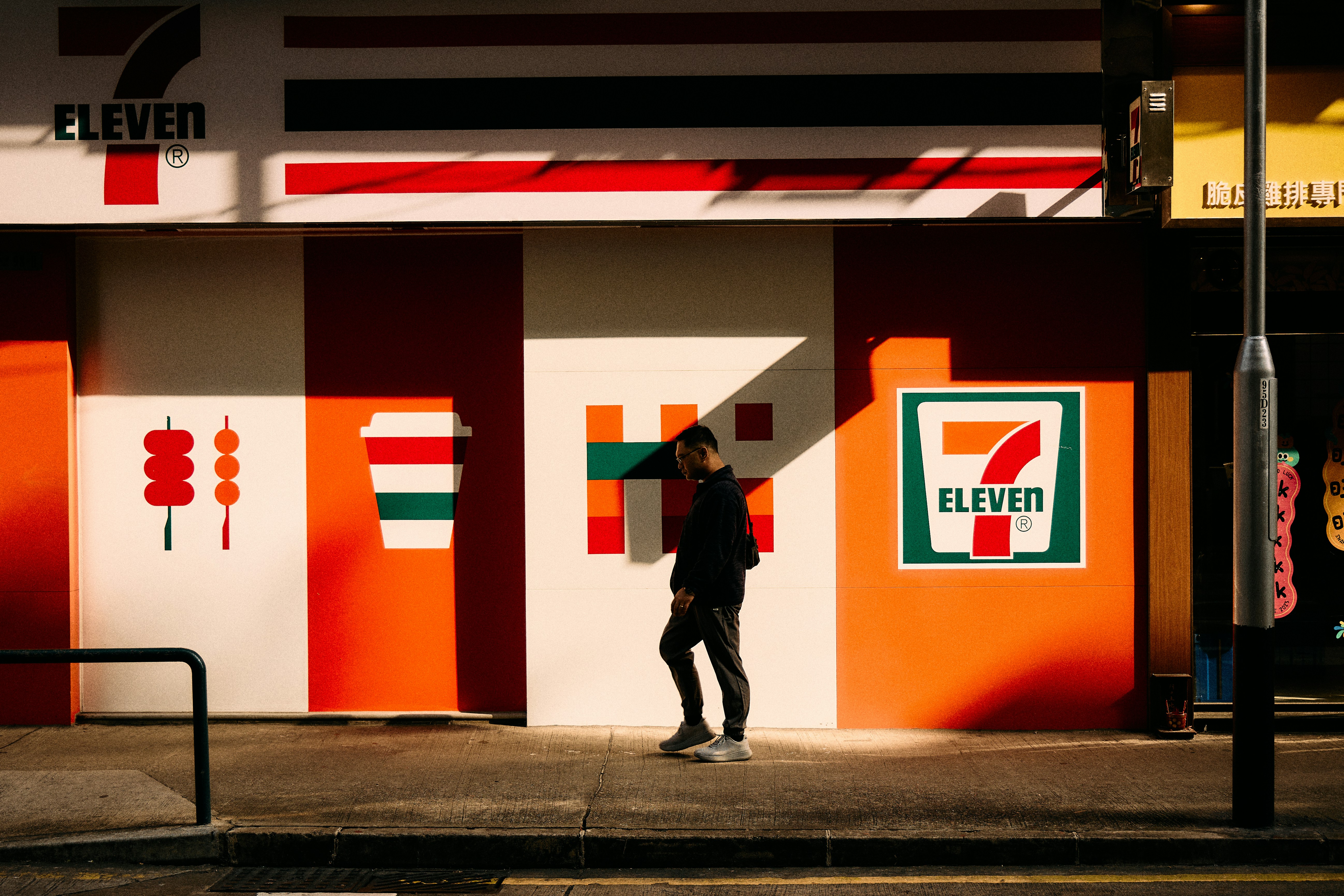 Man walking past a brightly colored 7-Eleven storefront with bold graphics in sunlight.