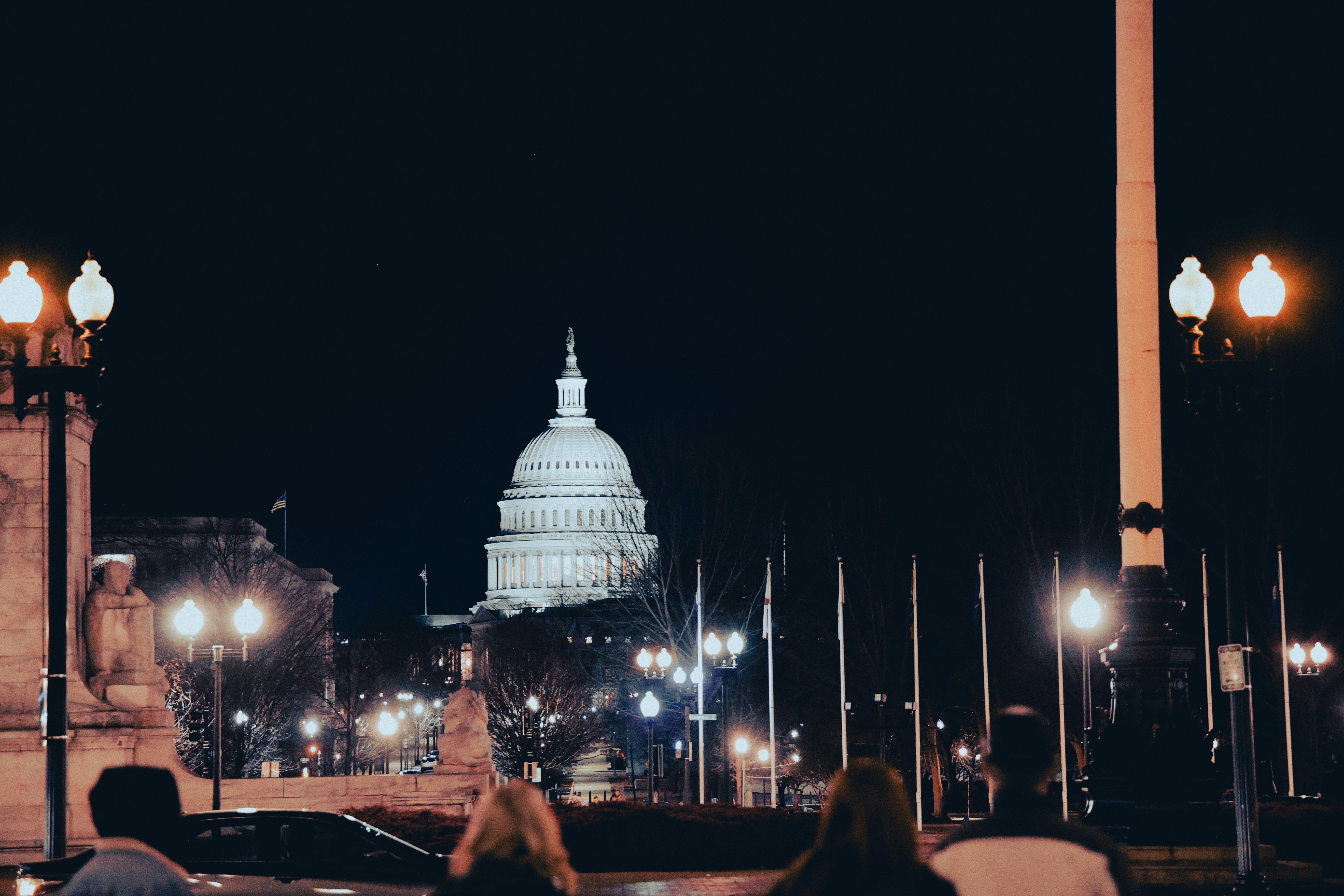 United States Capitol illuminated against the night sky, framed by glowing streetlights and statues with silhouetted pedestrians in the foreground.