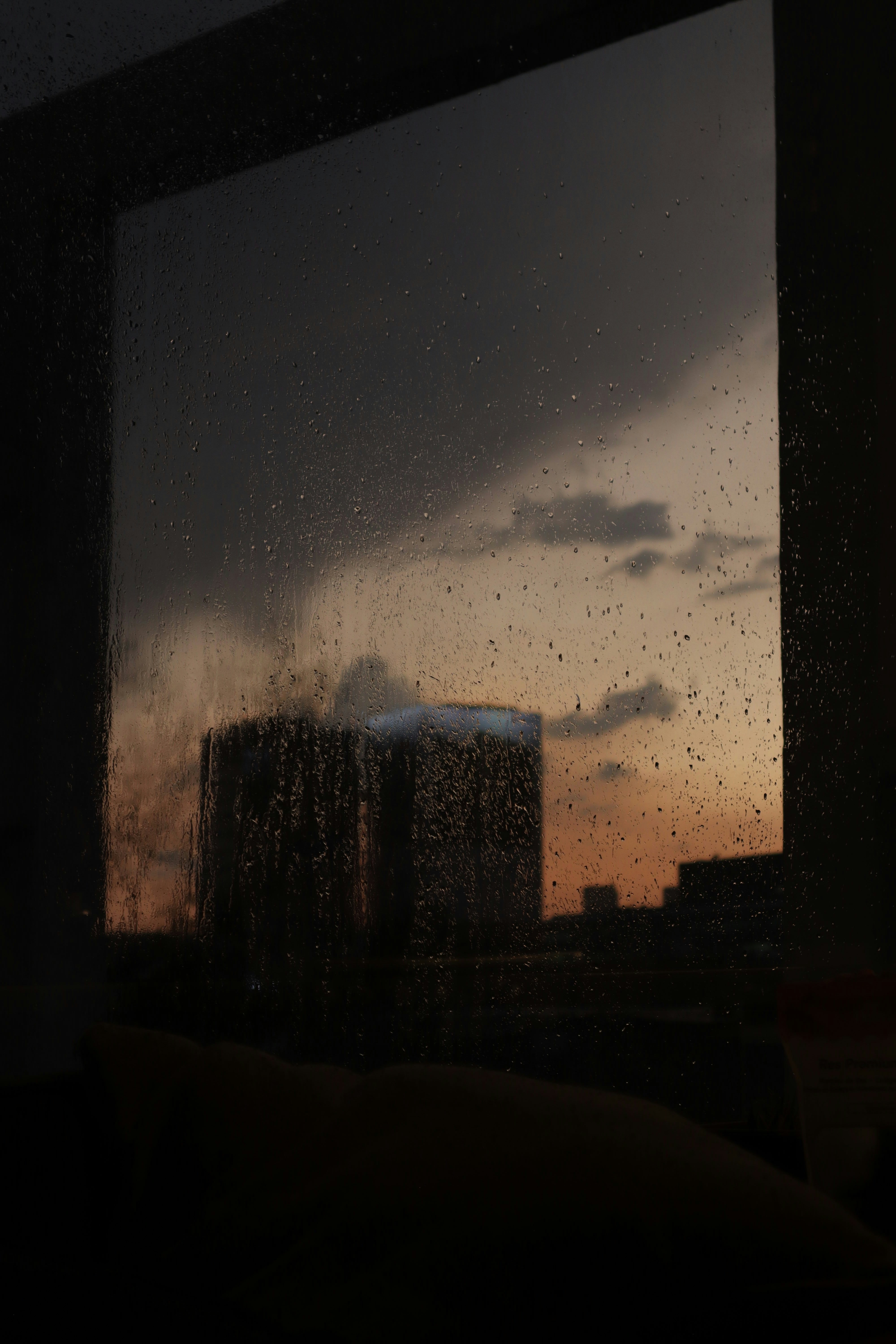 Raindrops on a window frame the silhouette of city buildings against a dusky sky, capturing the essence of an urban evening.