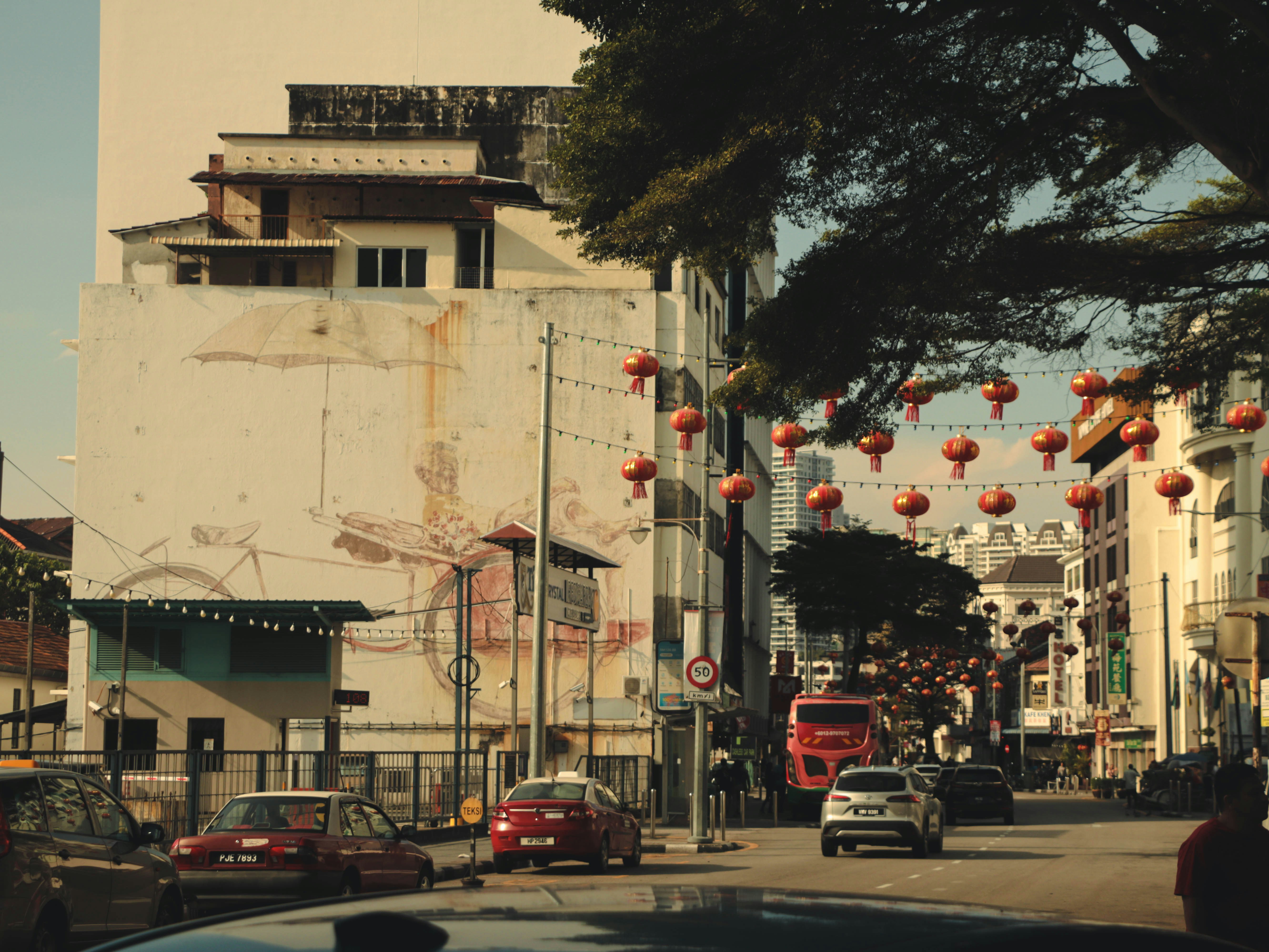 Urban street scene in Penang with a mural and hanging red lanterns under a tree.