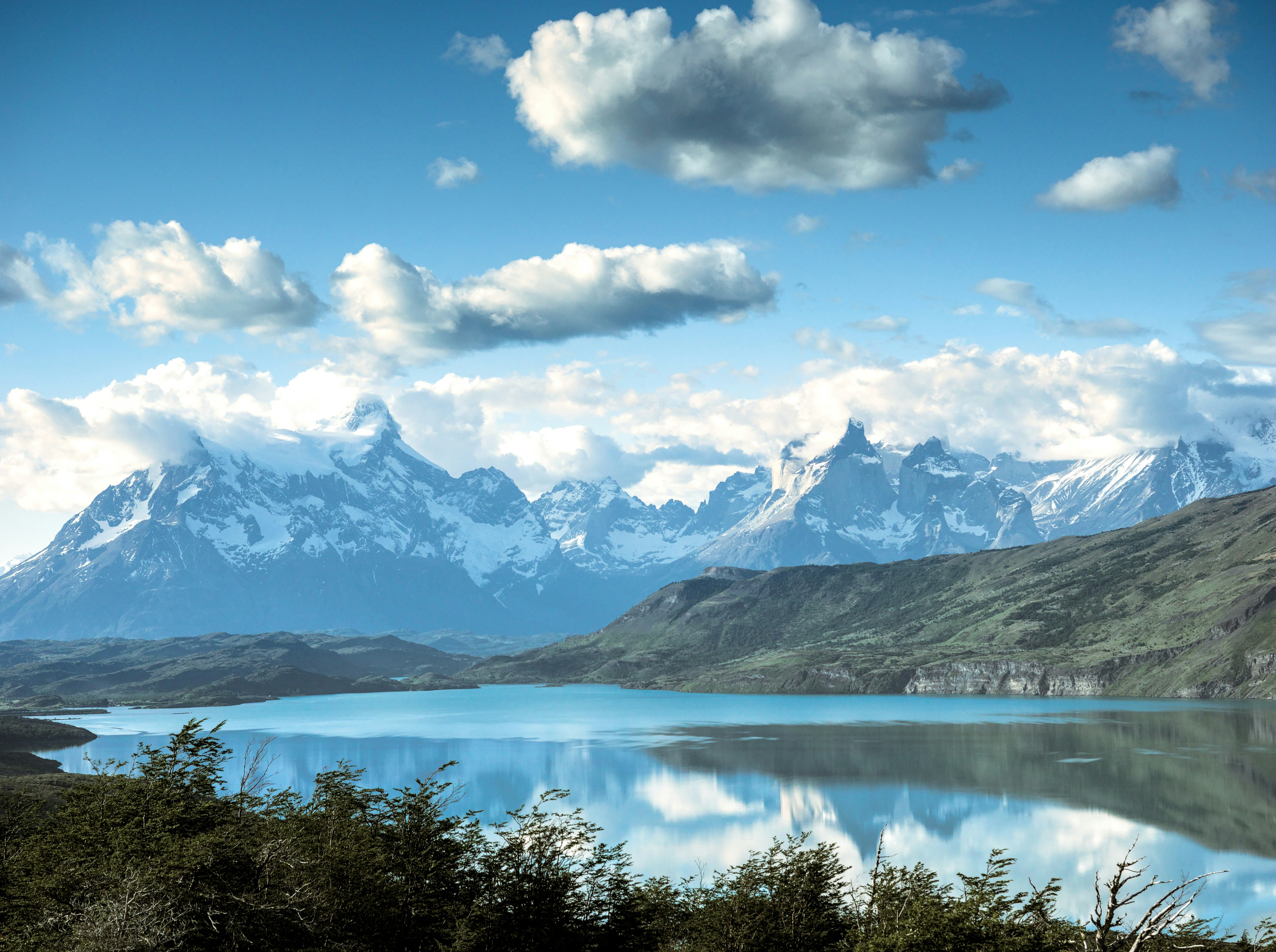 Snow-capped mountains and clouds reflected in a tranquil lake under a blue sky.