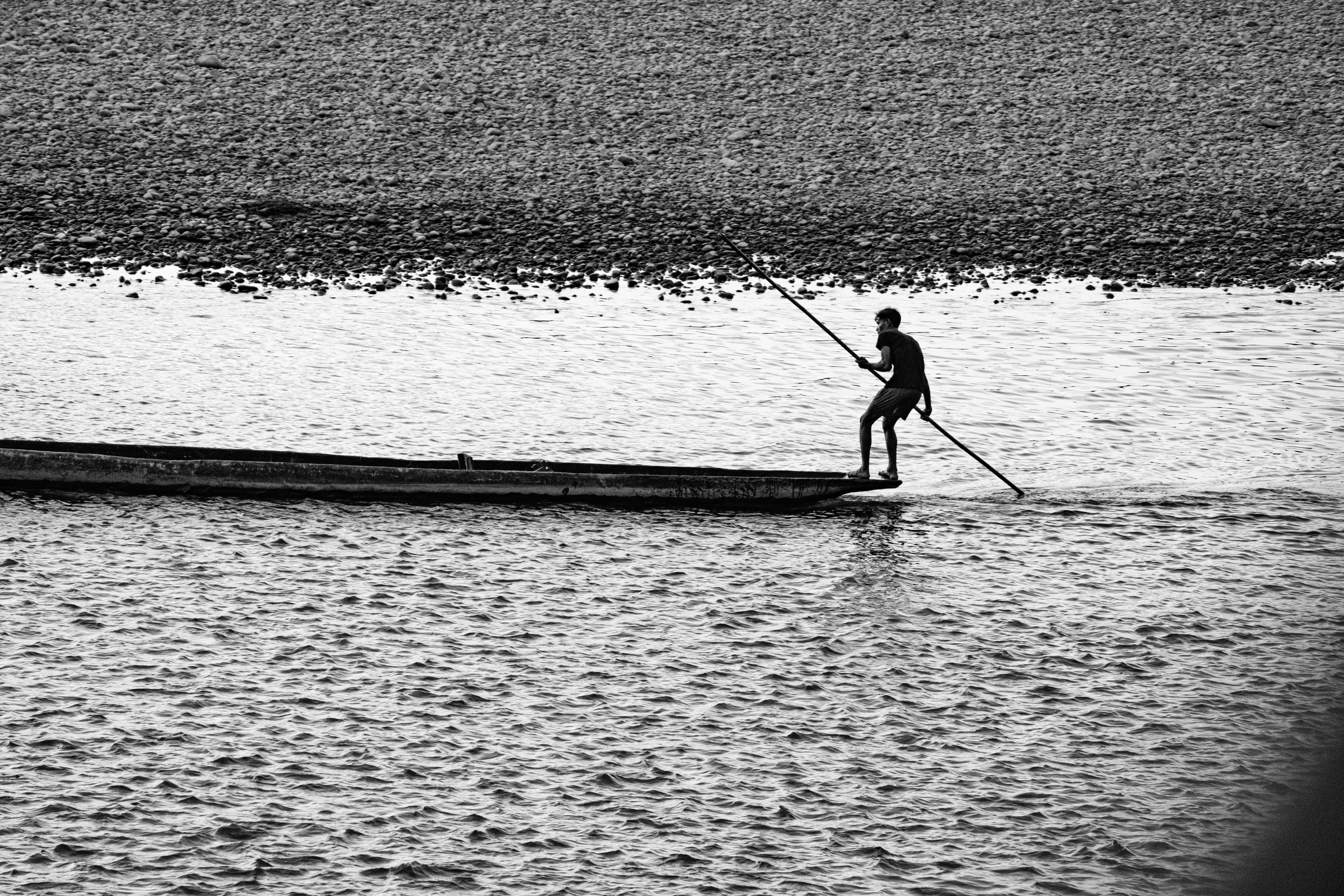Silhouetted figure navigating a narrow boat across a textured body of water with a rocky shoreline.