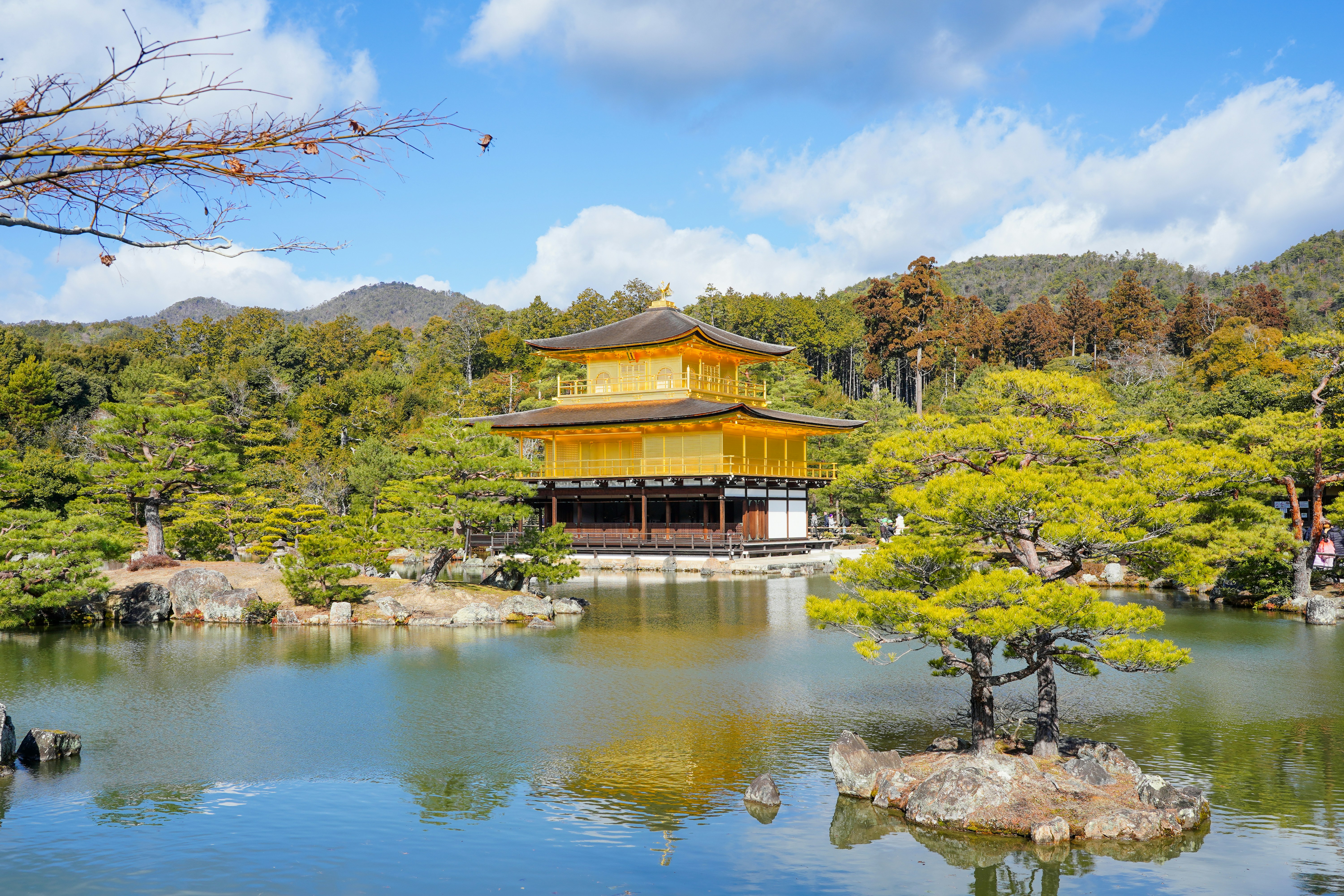Golden Pavilion reflects on the tranquil waters of a pond, surrounded by lush greenery under a bright sky.