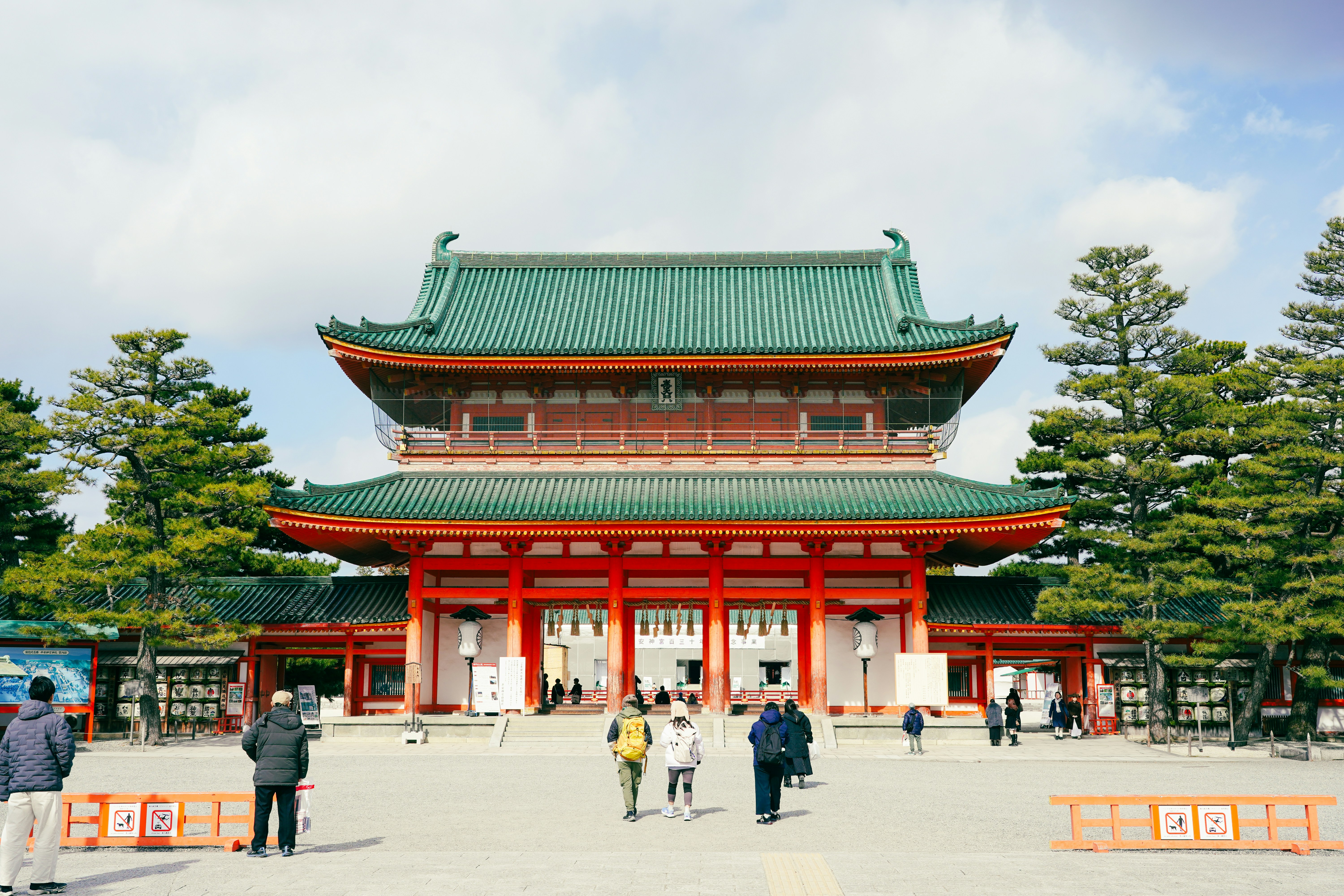 Vibrant torii gate of Heian Shrine with green-tiled roofs against a cloudy sky, surrounded by visitors and pine trees.