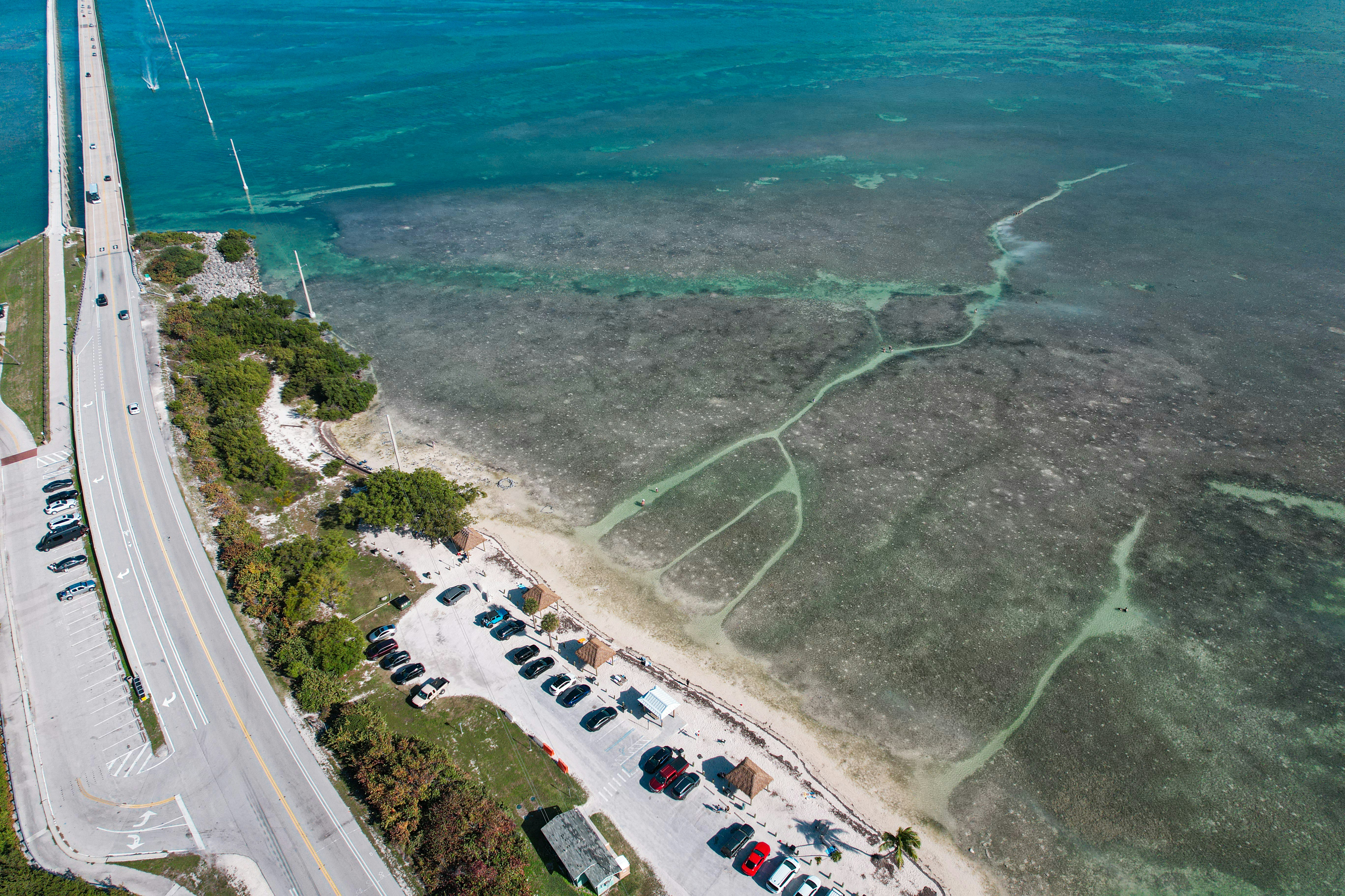 Aerial view of a bridge stretching over turquoise waters with cars parked along a sandy shore.
