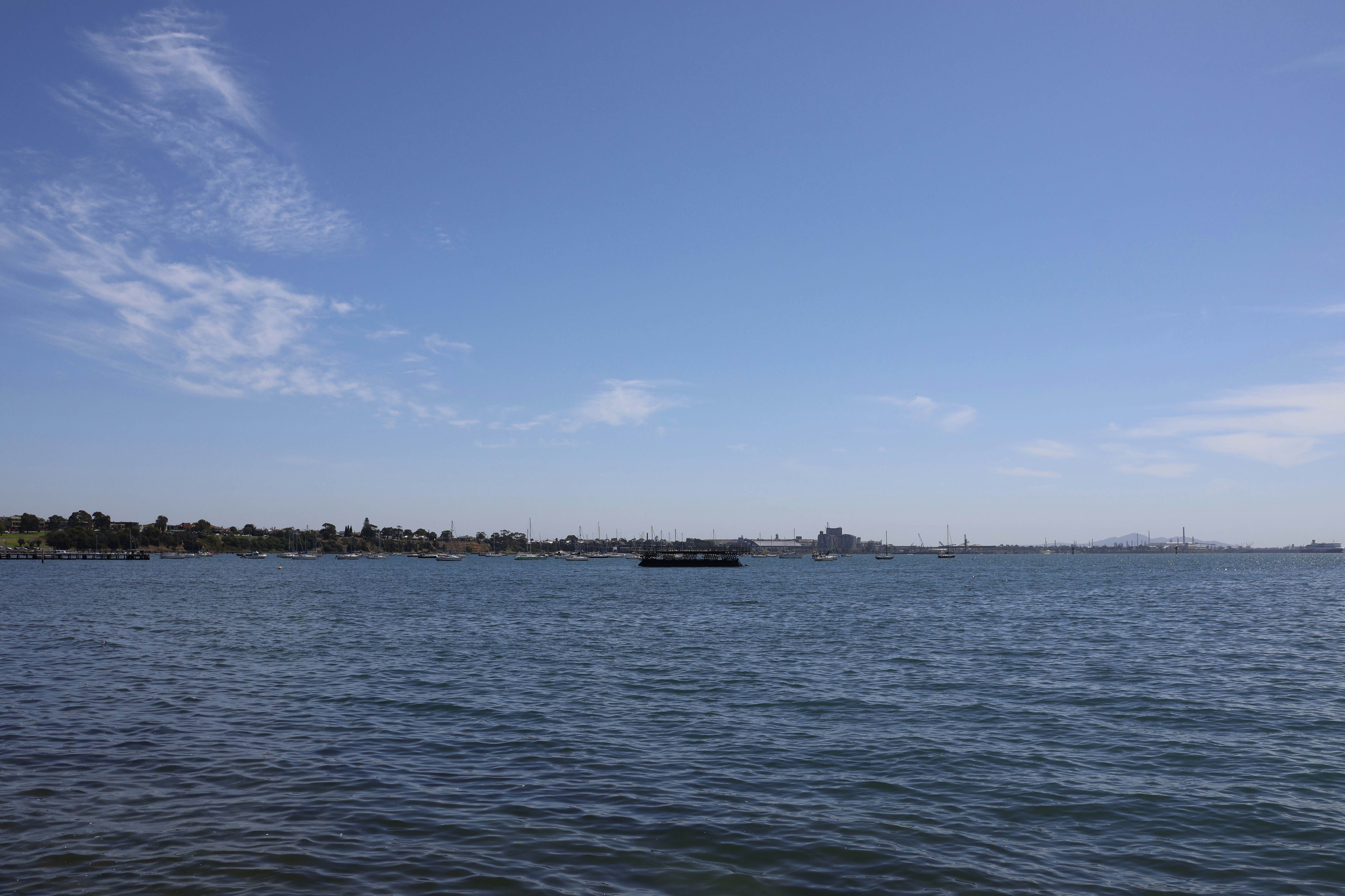 Calm waters with a distant city shoreline and scattered sailboats under a clear blue sky.