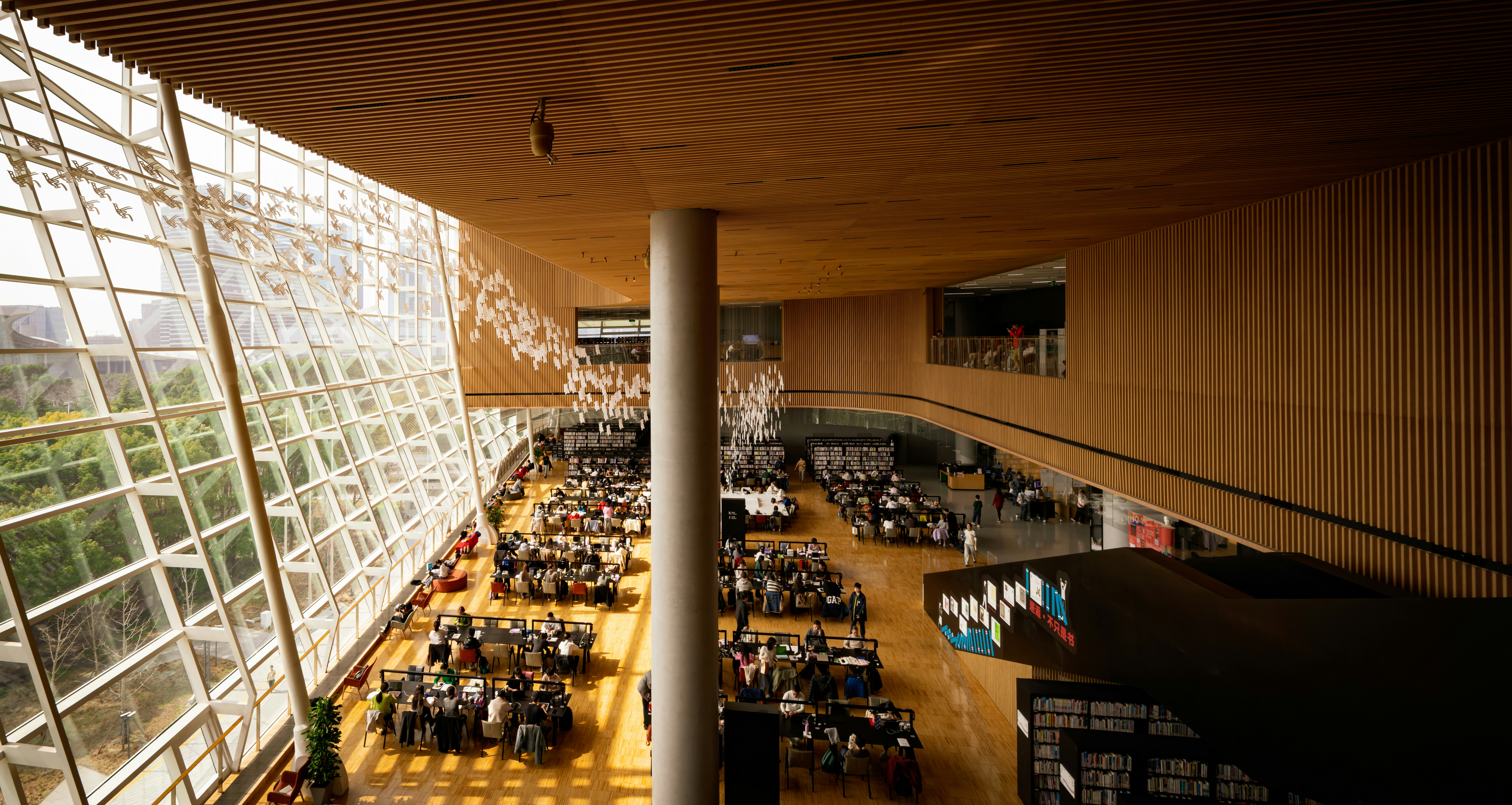 Spacious library interior with large windows and wooden accents, bustling with people at study tables.