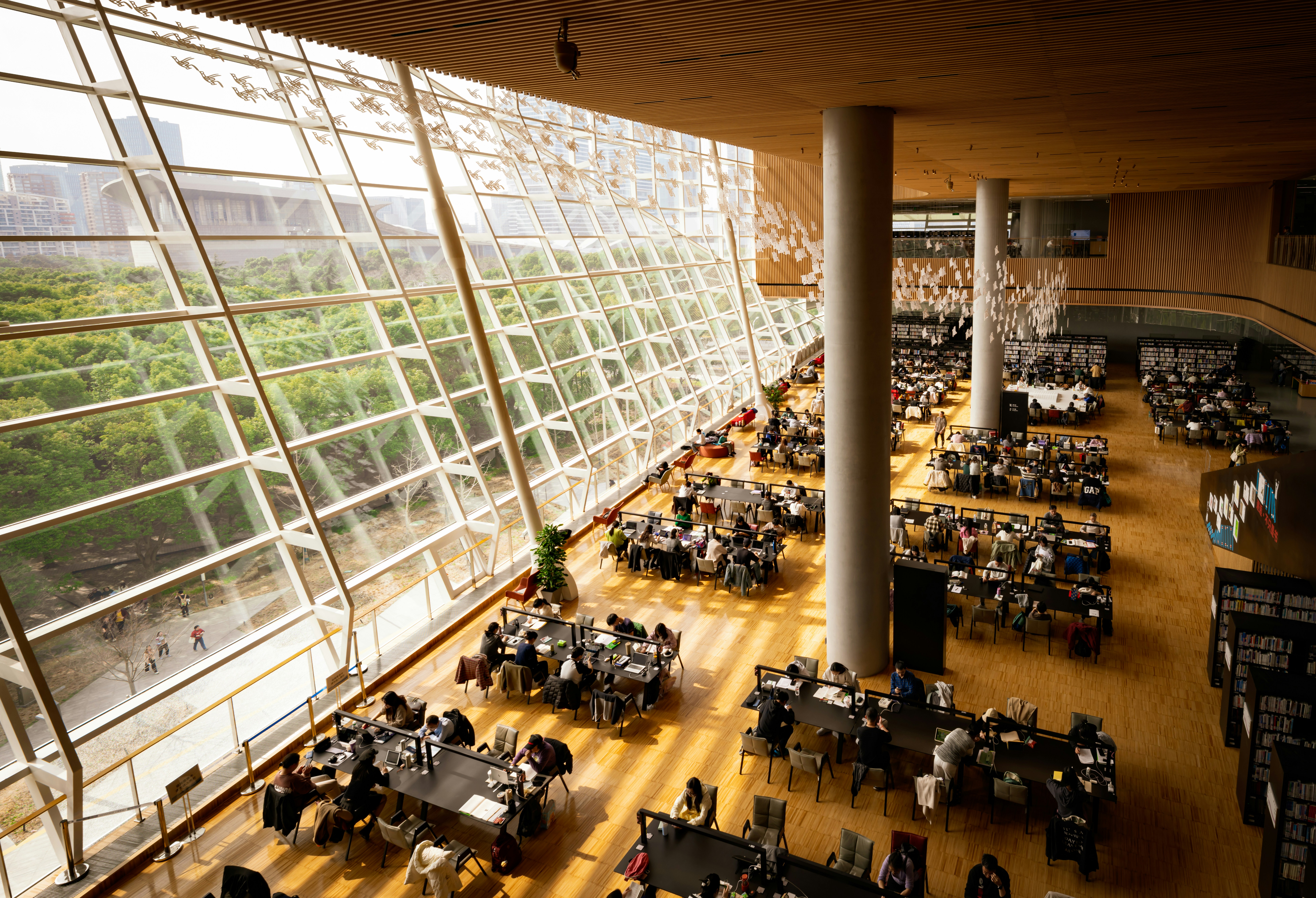 Spacious library interior filled with natural light through large angled windows, bustling with people at workstations.