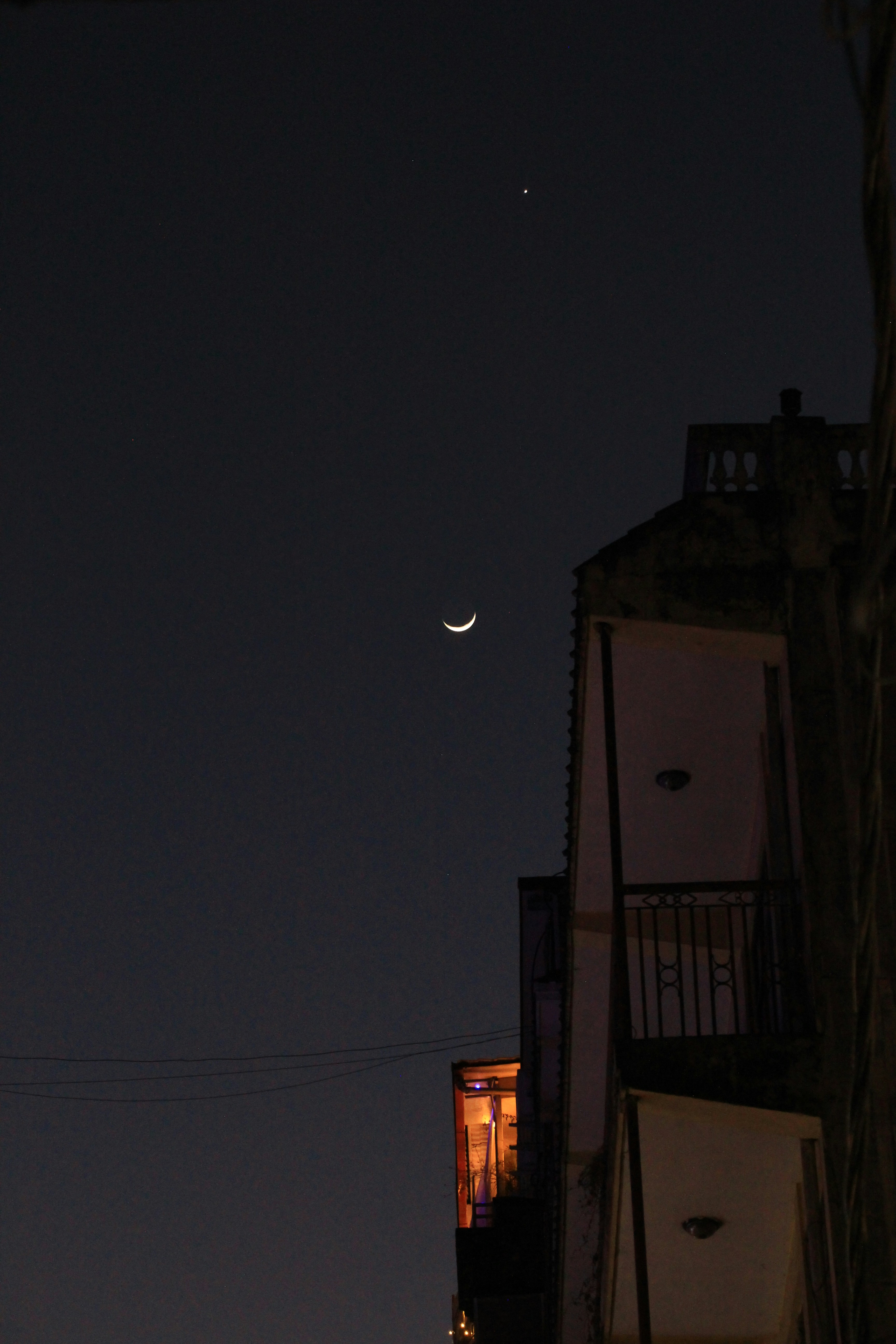Crescent Moon Over a Dim Urban Facade with a Warm Balcony Glow