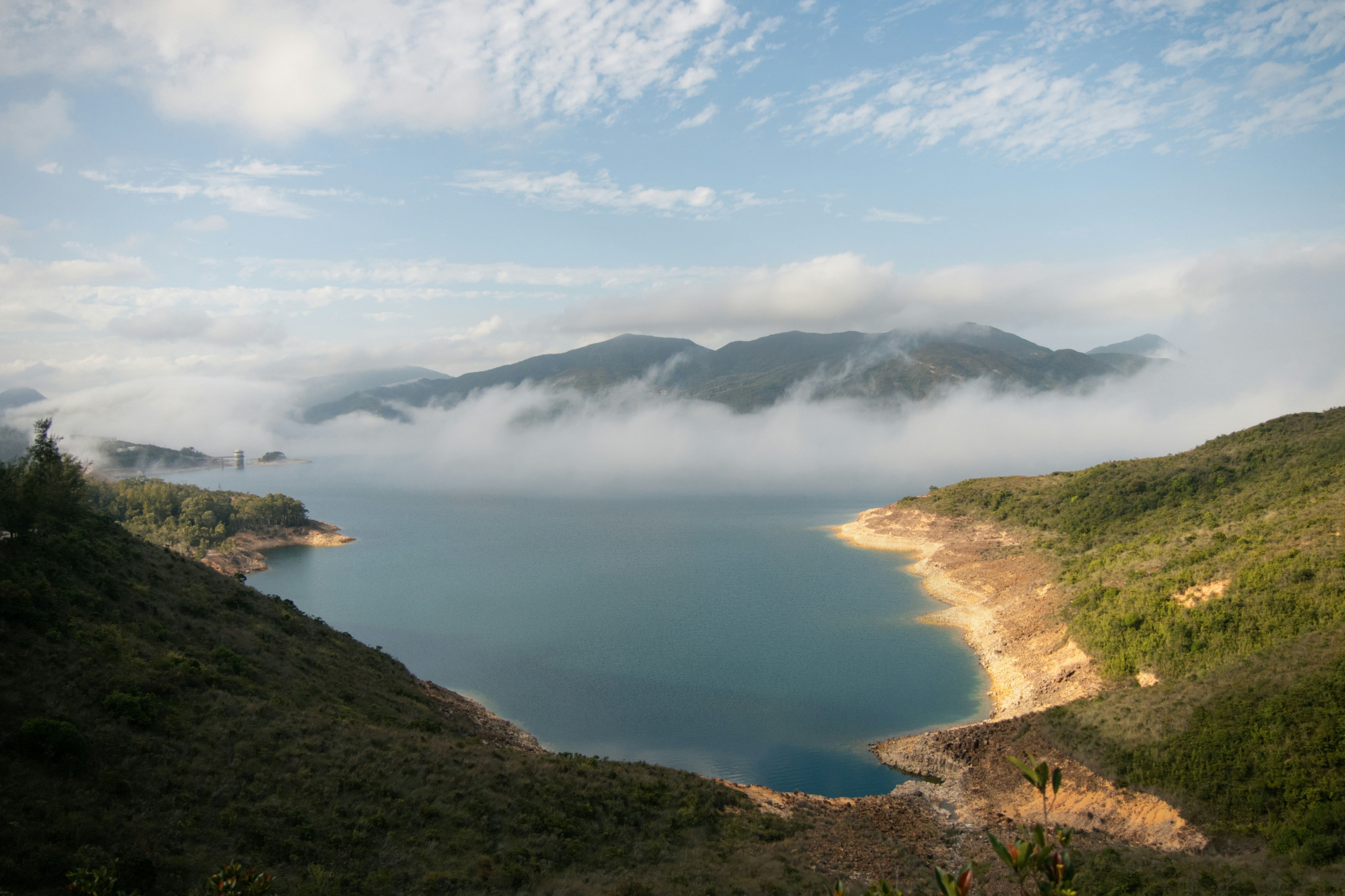 Tranquil lake bordered by lush hills and veiled in low-lying mist under a partly cloudy sky.